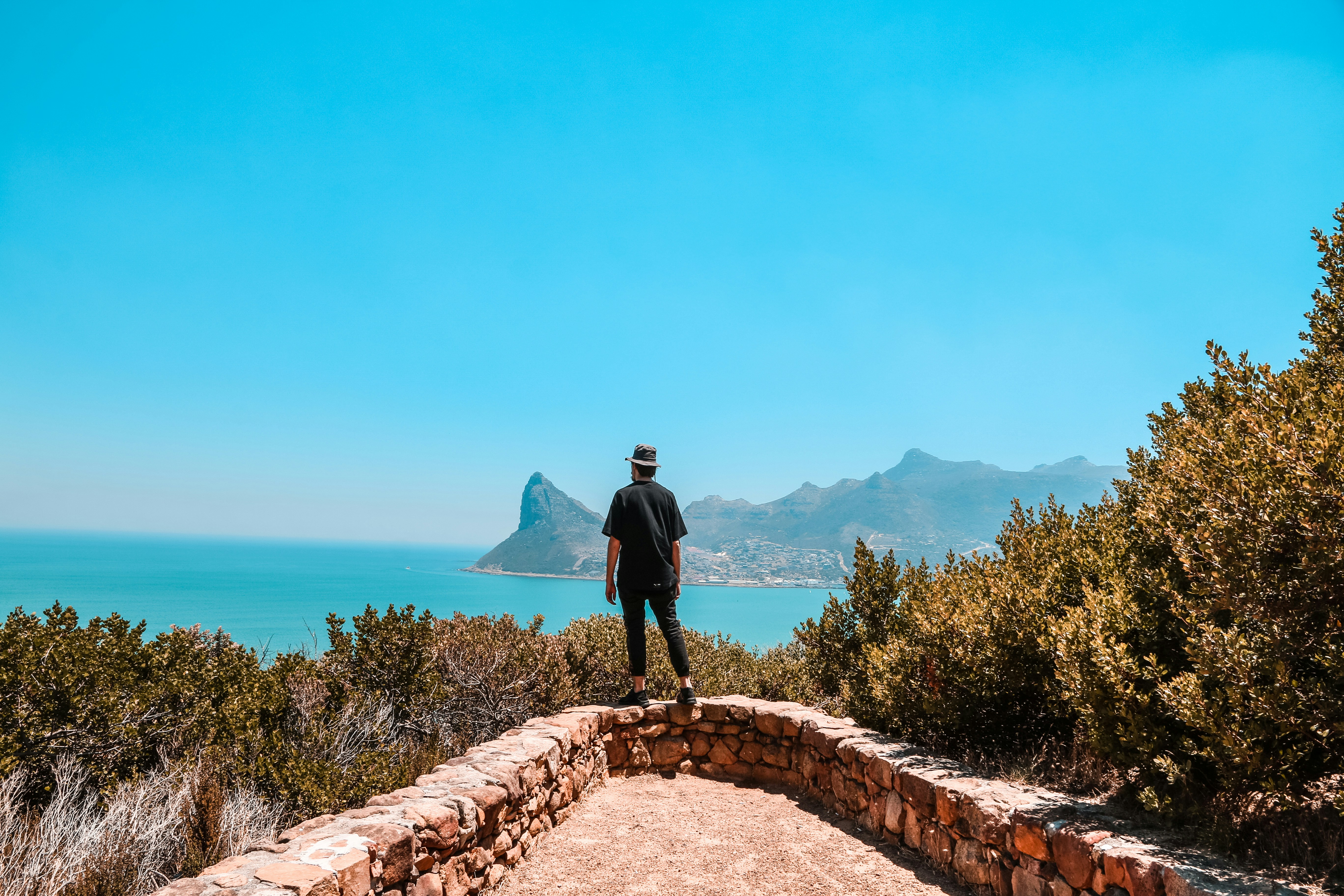 Individual standing on a stone path overlooking a serene coastal landscape with mountains in the background.