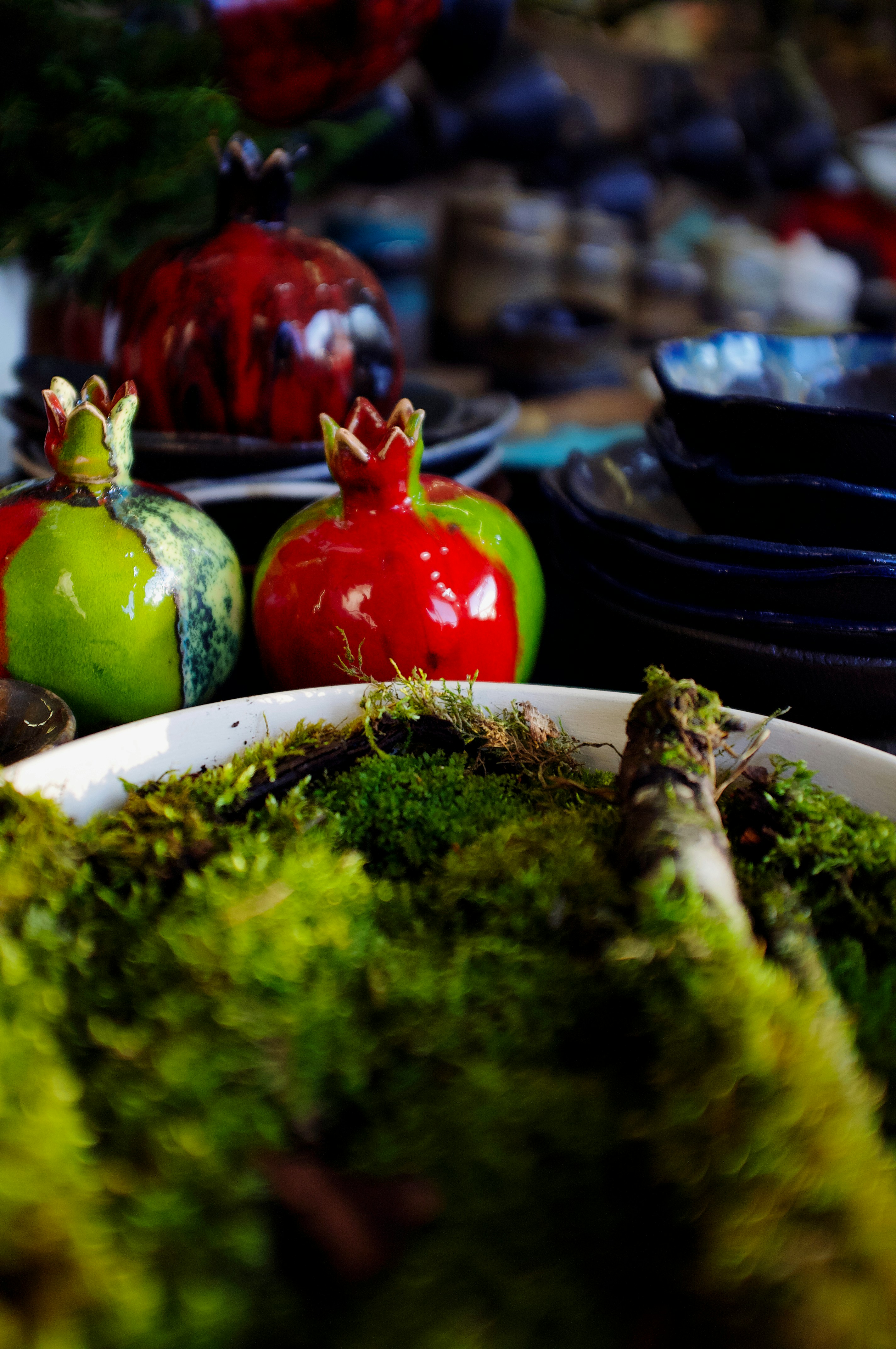 Colorful ceramic fruits nestled in lush green moss, creating a vibrant contrast with the surrounding pottery. The scene evokes a playful harmony between art and nature.