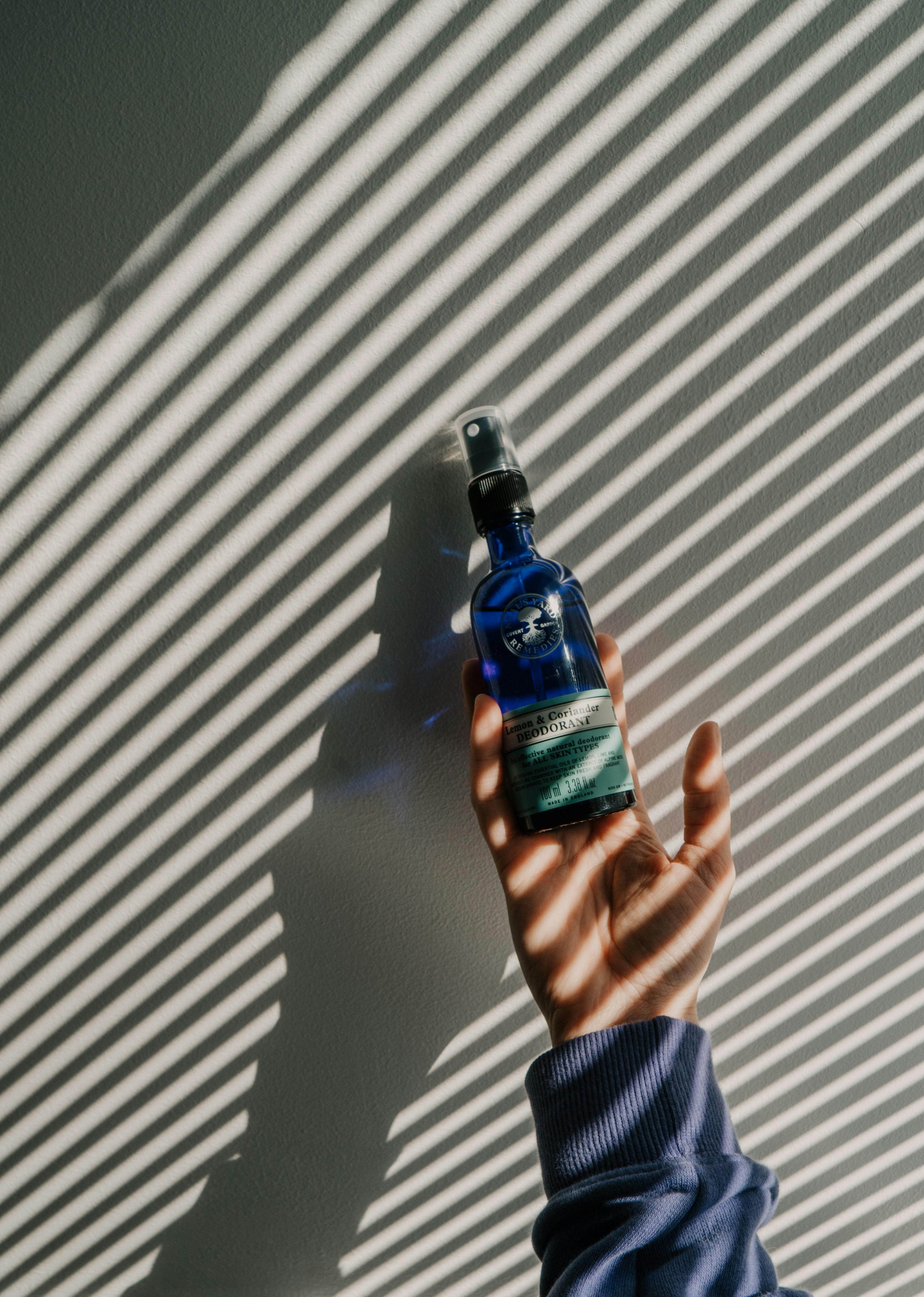 A hand holds a blue spray bottle against a backdrop of striking diagonal shadows. The interplay of light and shadow creates a dynamic visual effect.