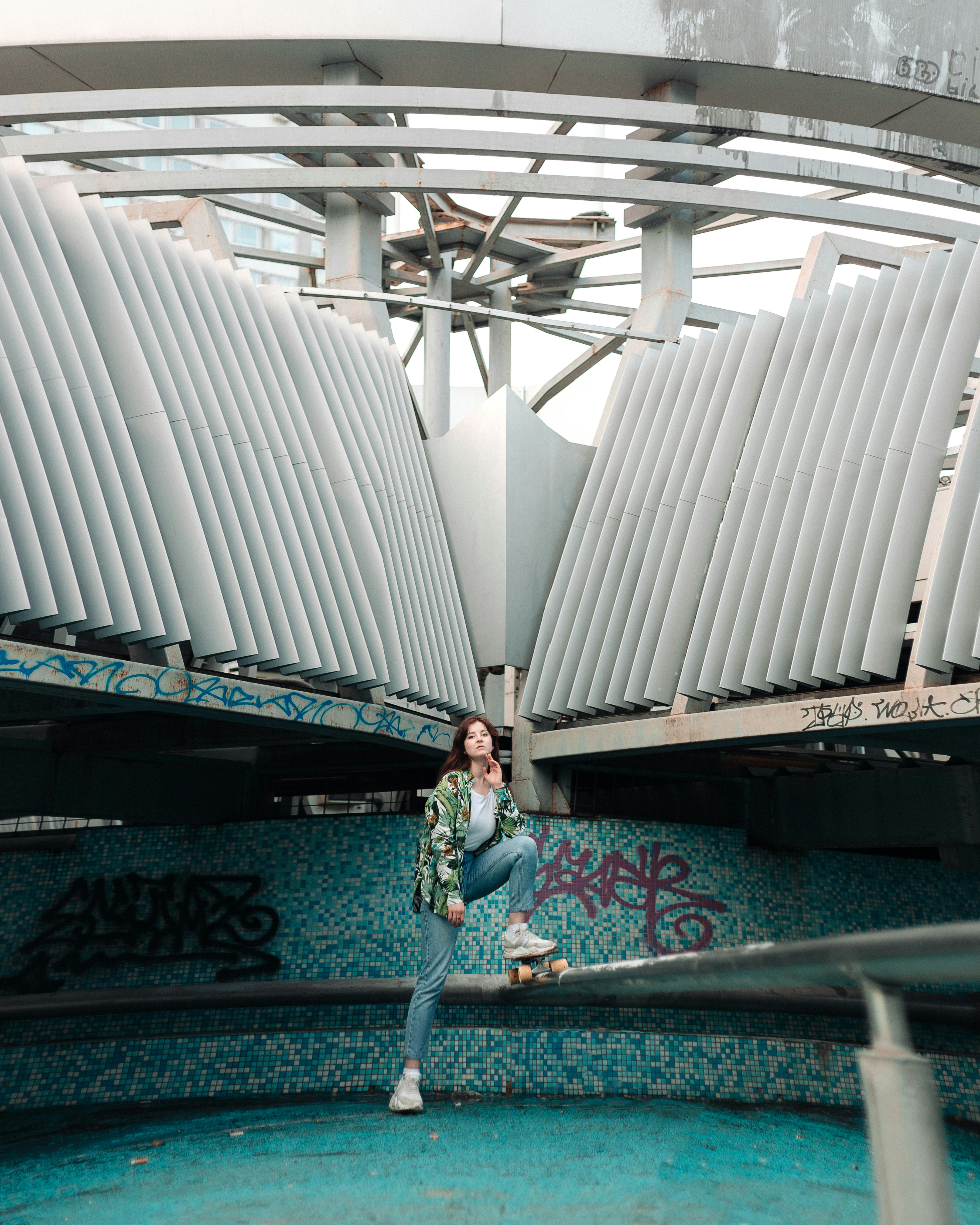 A young woman poses confidently on a railing beneath a striking architectural structure, surrounded by vibrant graffiti and tiled patterns.