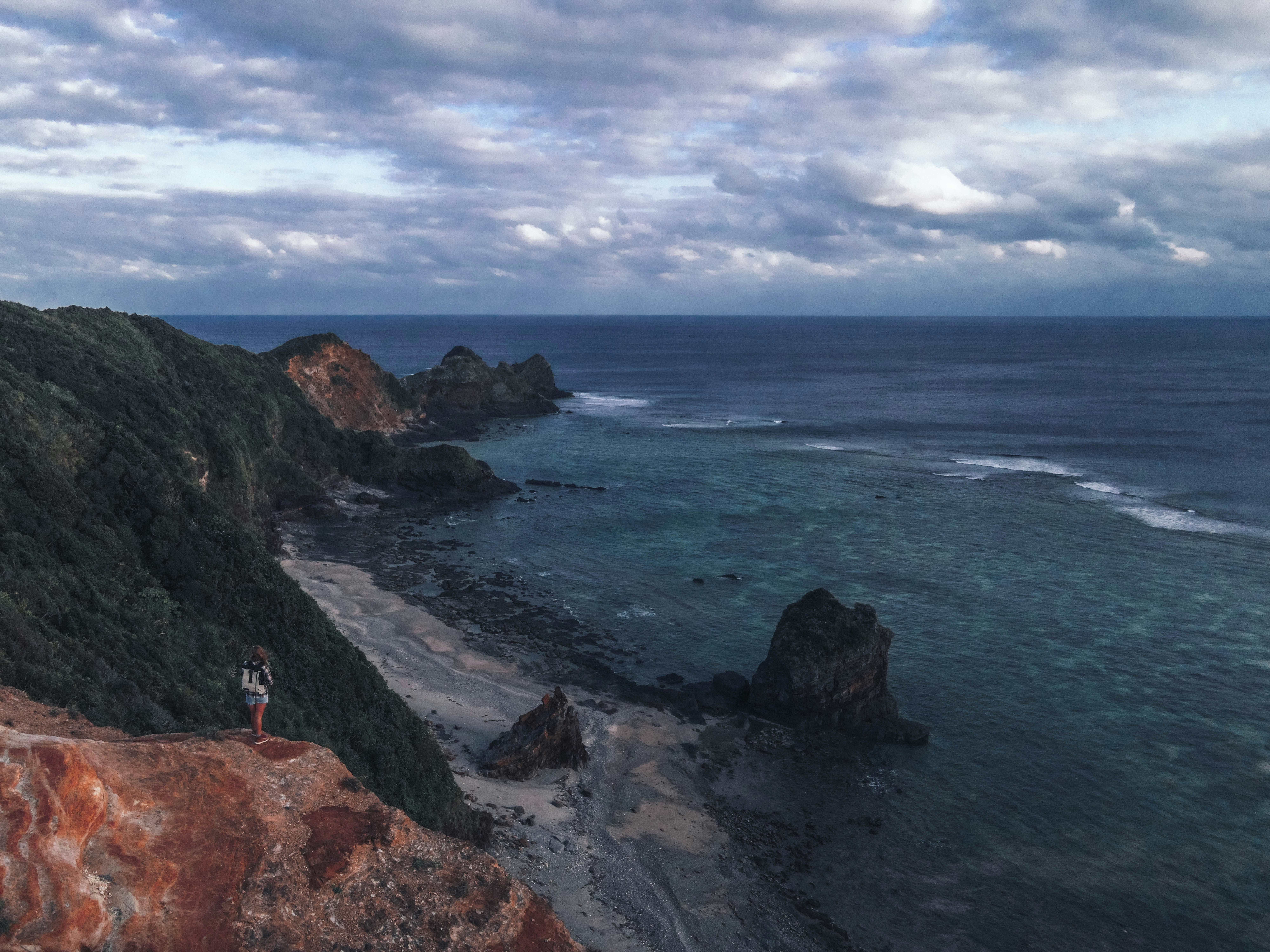 Individual standing on rocky cliff overlooking expansive ocean and rugged coastline under a cloudy sky.