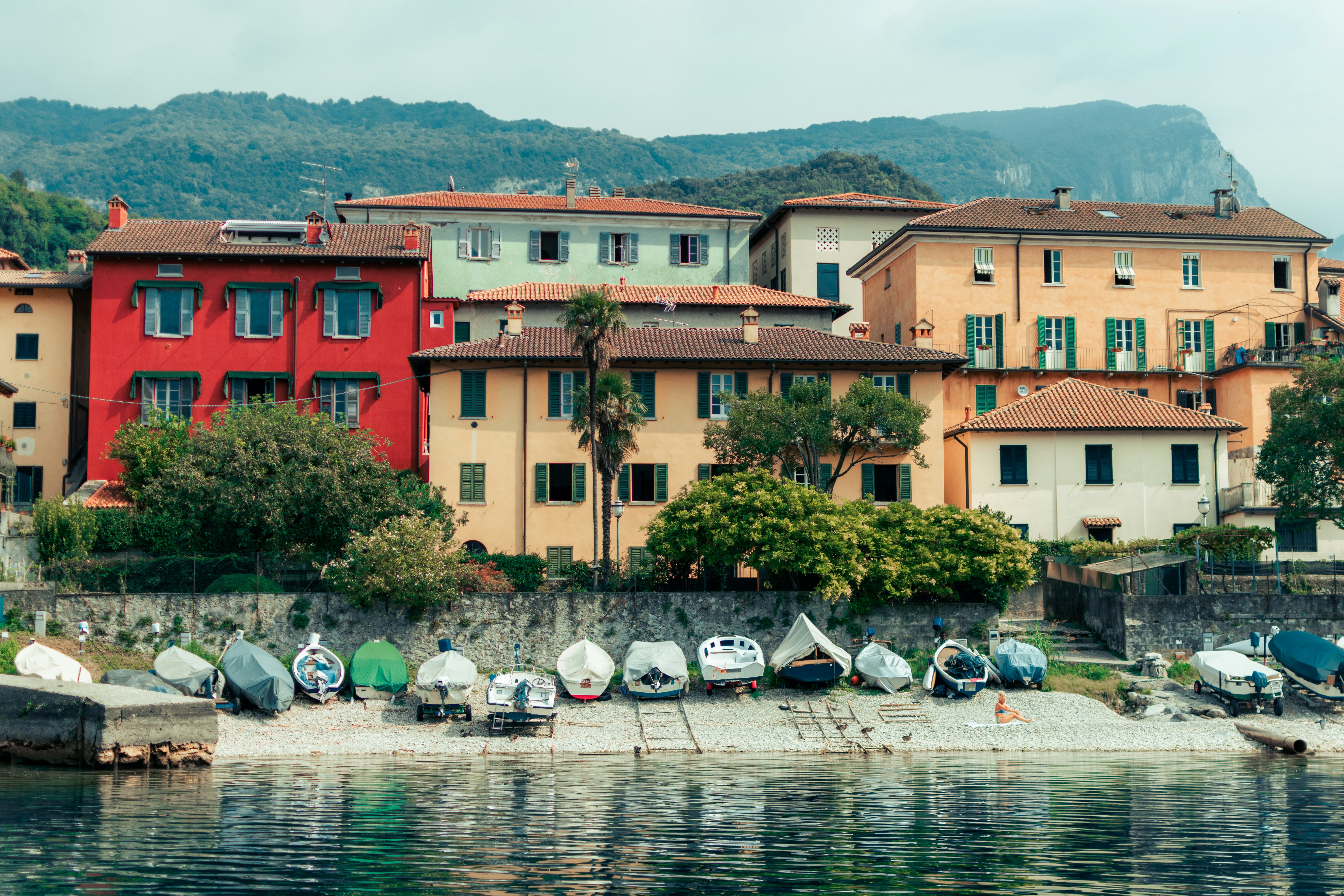 Colorful houses lining a serene lakeside, with boats resting on a pebbled shore. The scene captures the essence of tranquil village life.