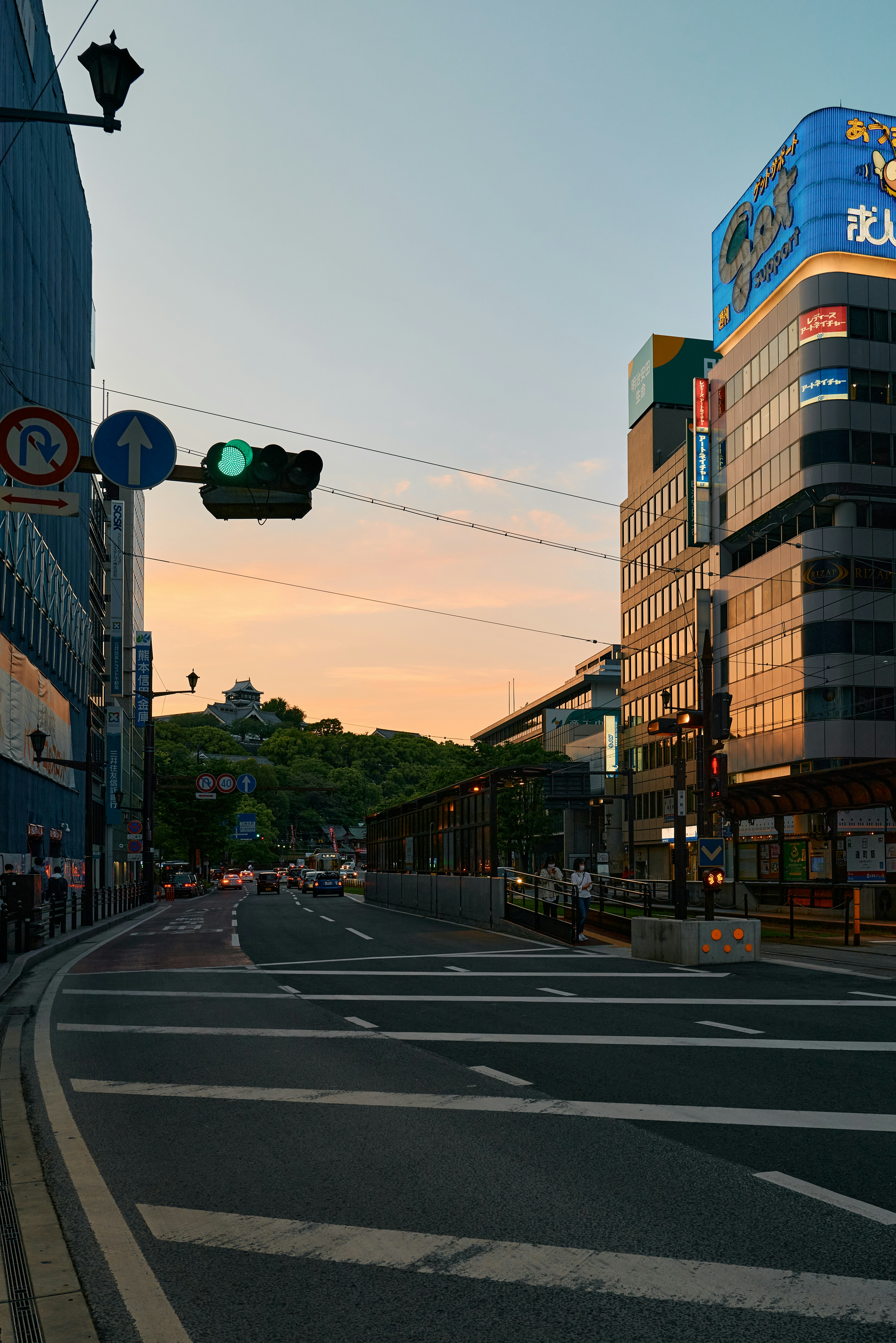 A bustling city intersection illuminated by traffic lights, with a distant castle silhouette against a pastel sunset sky.