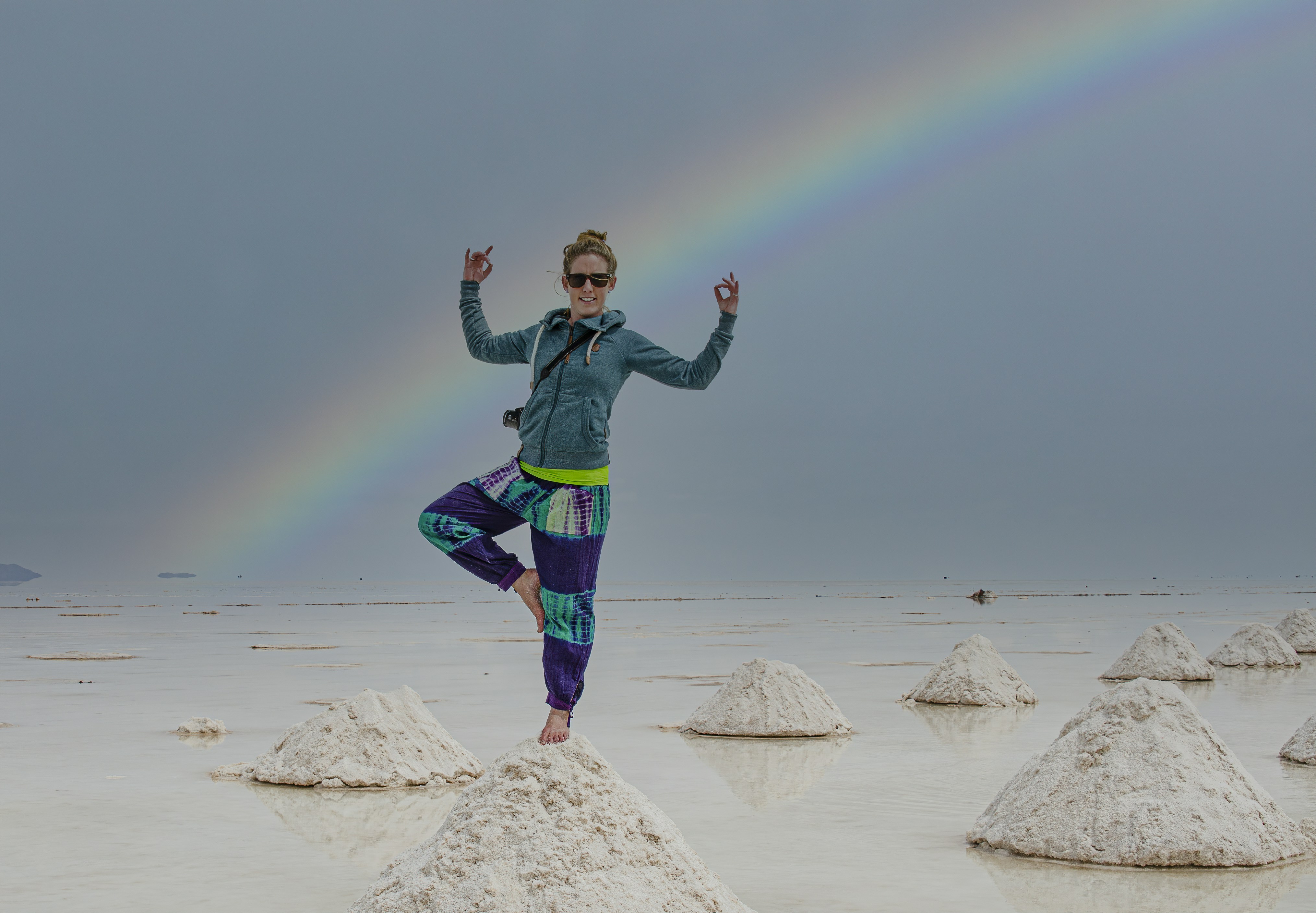 A person balances on a salt mound in a vast salt flat, with a vibrant rainbow arching across a cloudy sky.