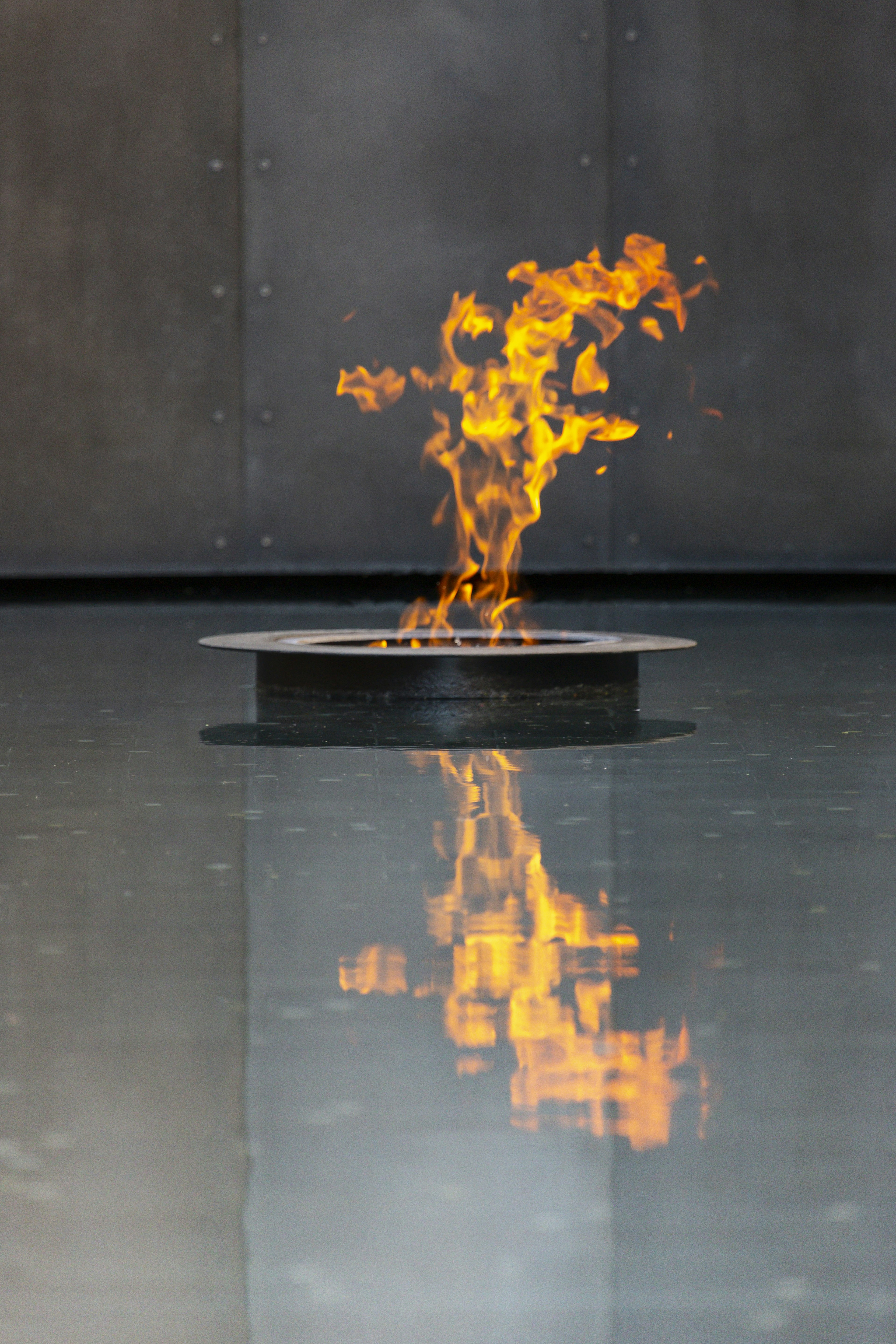 Flame flickering above a circular black base, mirrored on a glossy surface, creating a mesmerizing visual effect.