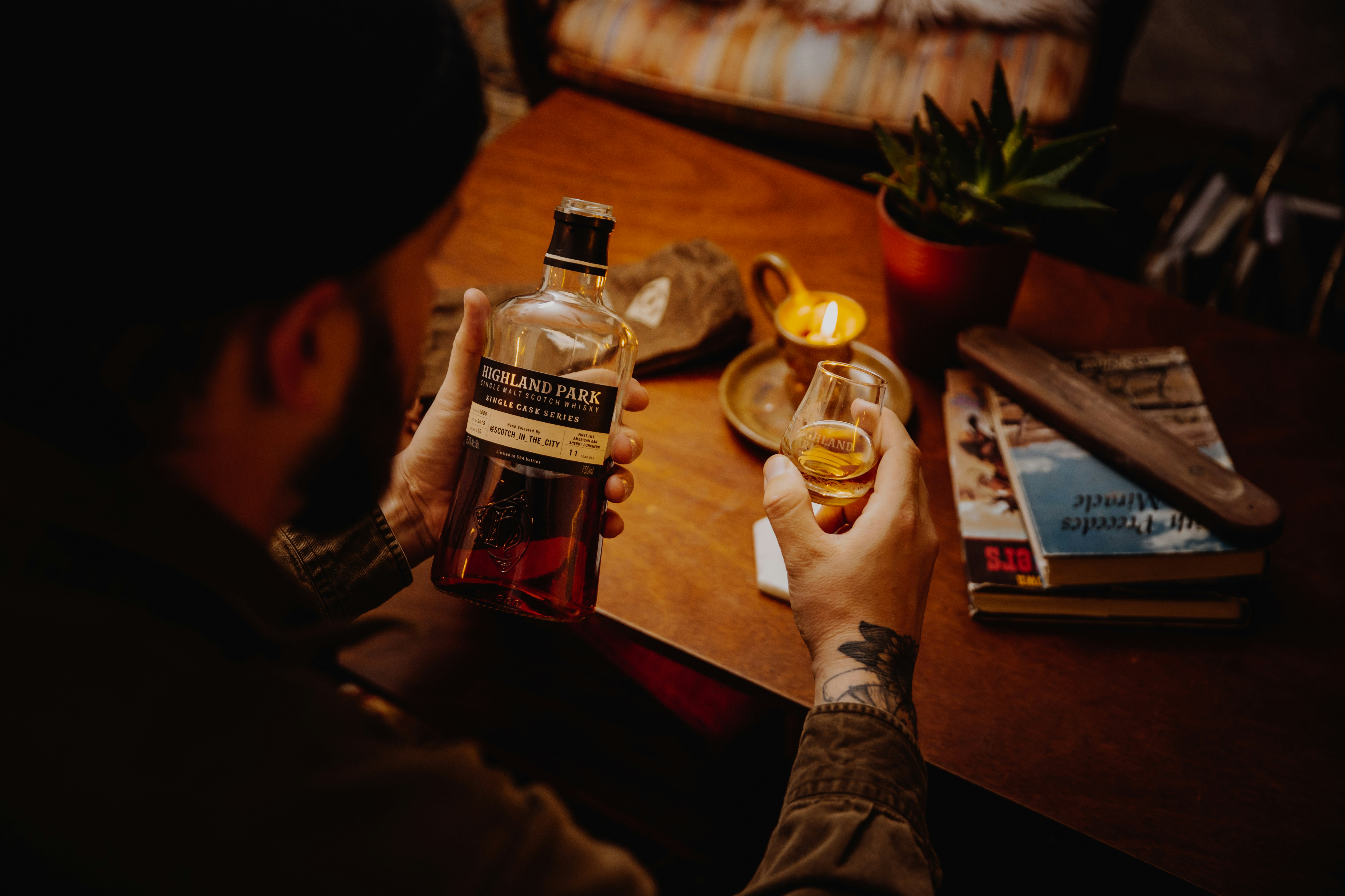 Man holding a bottle and glass of whiskey in a cozy, dimly lit room with books and a candle on the table.