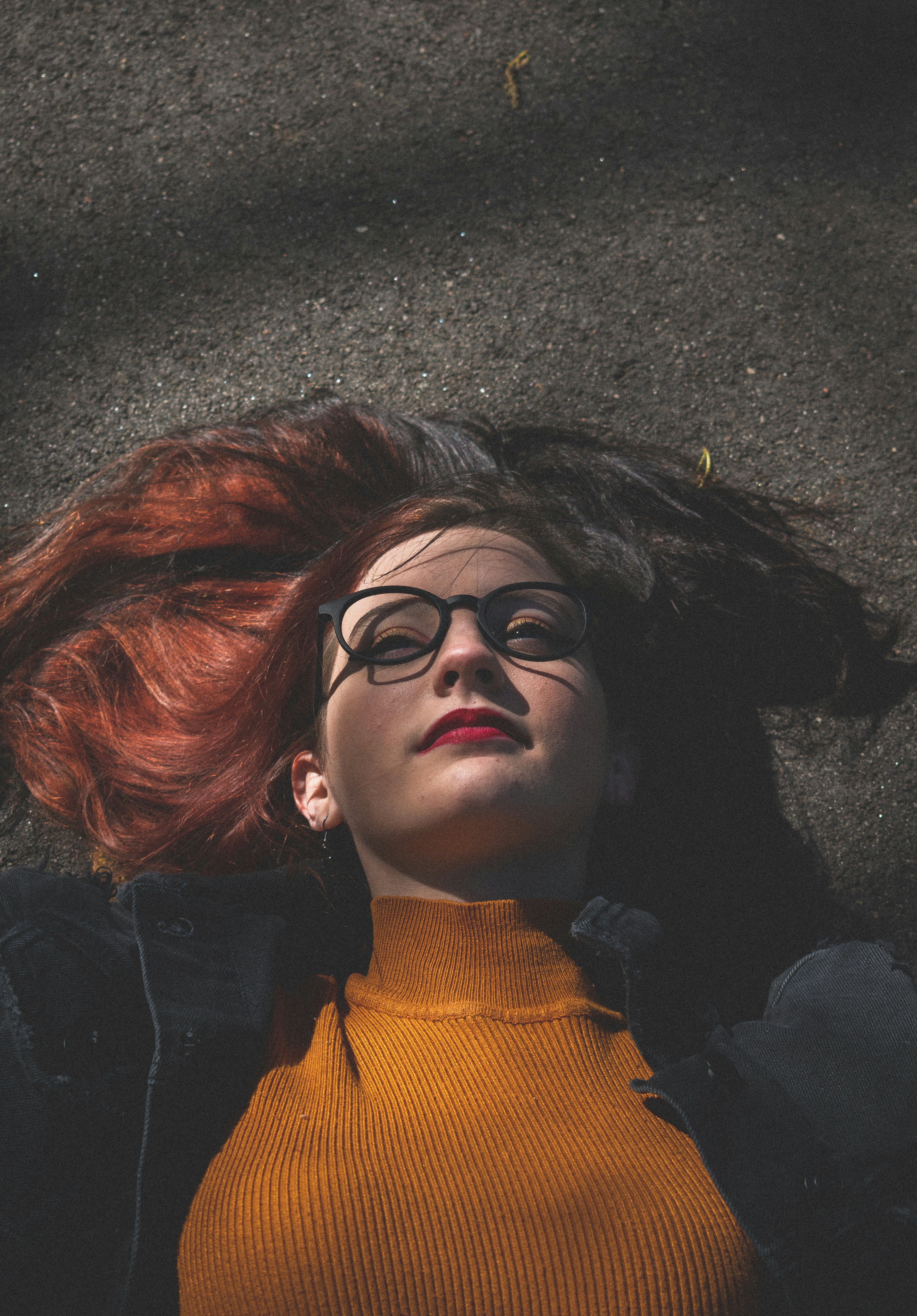 A young woman with vibrant red hair and glasses lies on a textured surface, exuding a sense of calm and introspection.