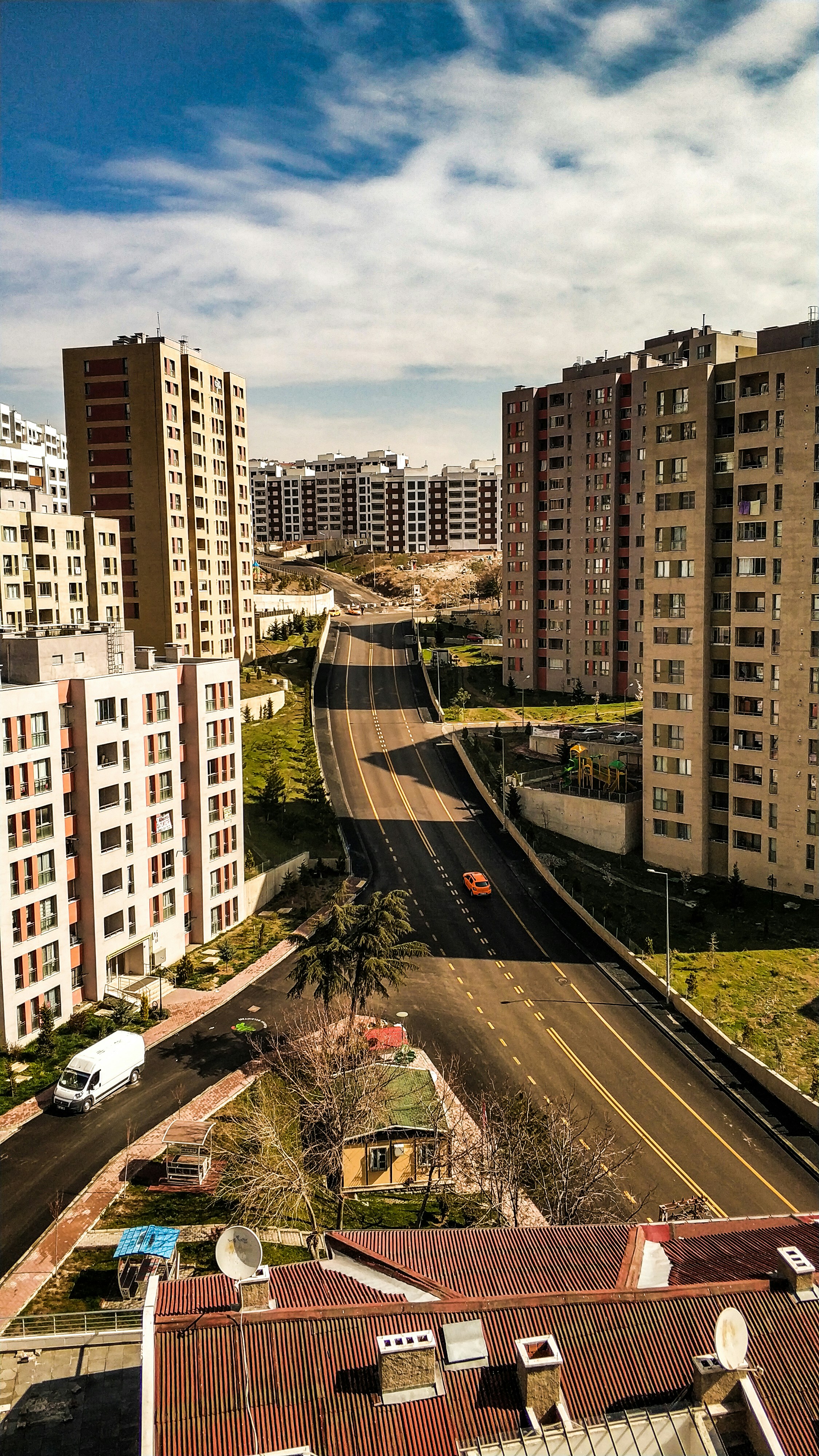 A winding road cuts through a contemporary urban landscape, flanked by tall residential buildings and patches of greenery.
