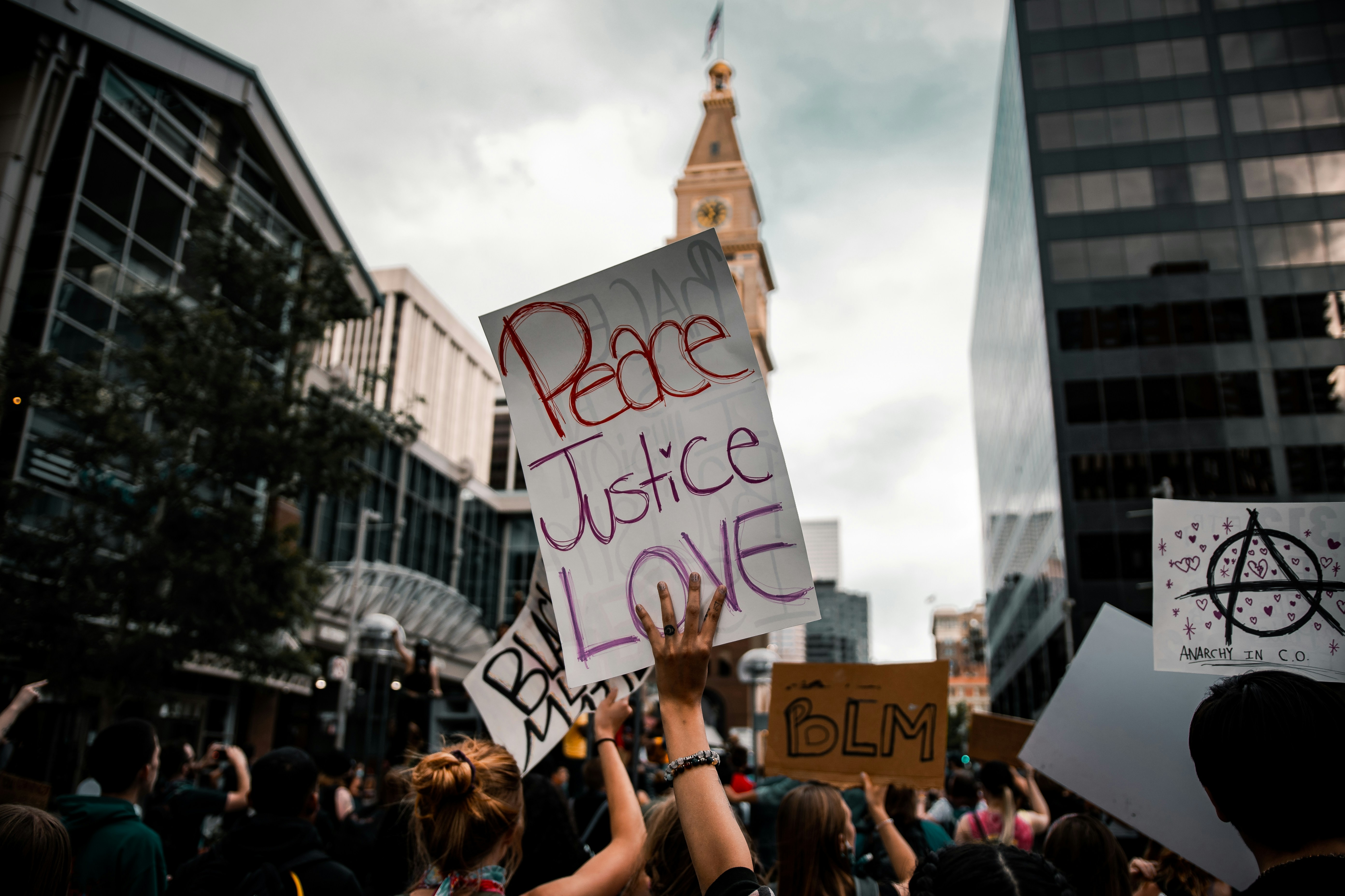 A diverse crowd holds up signs advocating for peace, justice, and love during a protest in an urban setting.