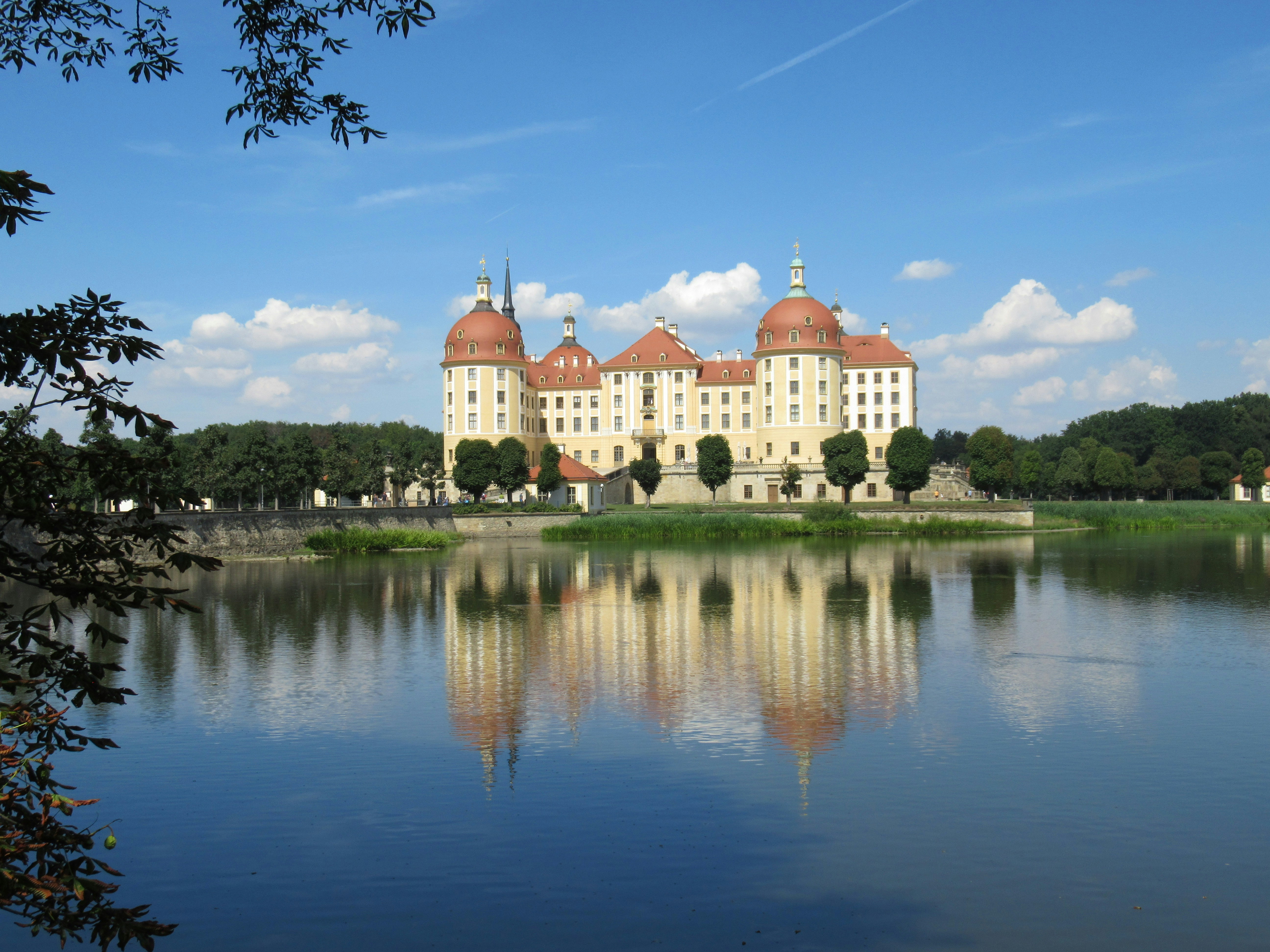 Castle-like mansion with red-domed towers rises across a calm lake, its reflection perfectly mirrored in the water beneath a bright, cloud-dotted sky.