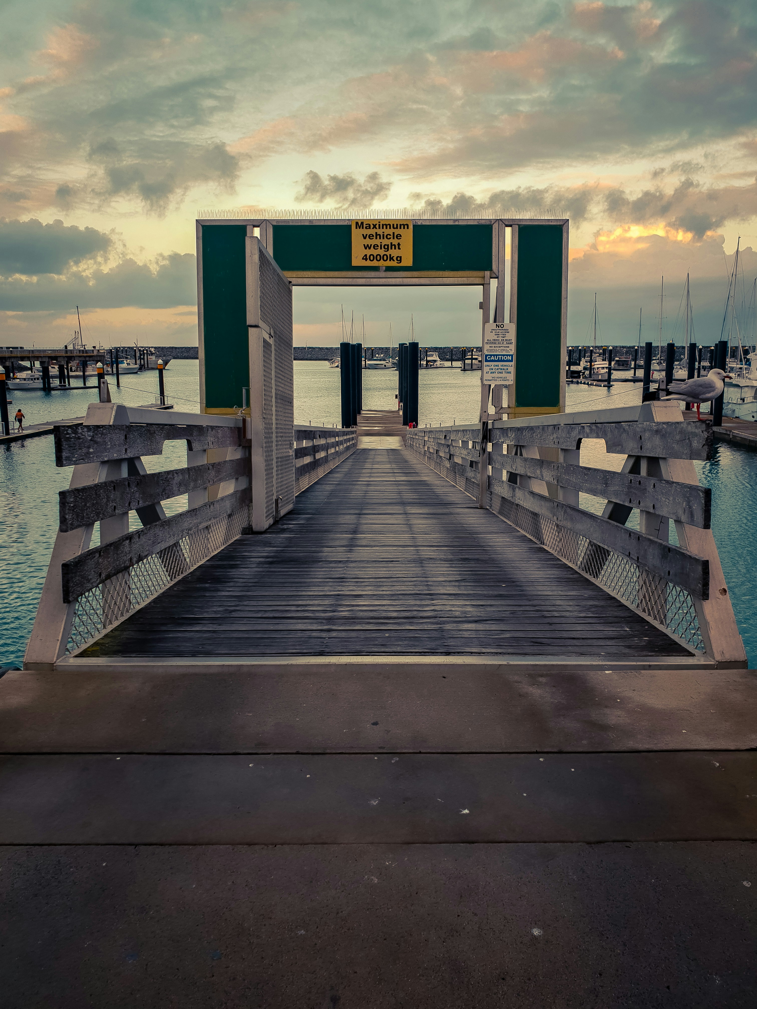 Wooden dock leading to a marina at sunset, framed by a green archway marked with weight limits. Calm waters reflect the evening sky.