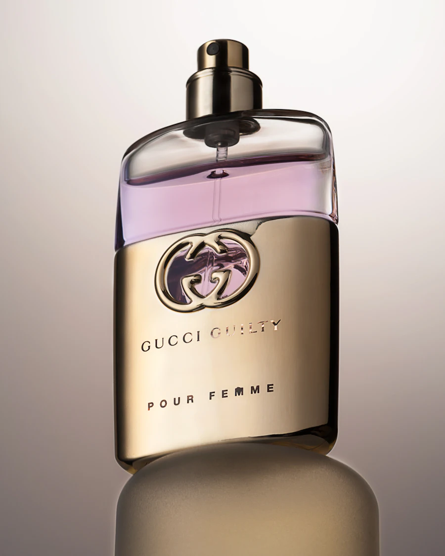 Gold-toned Gucci Guilty Pour Femme fragrance bottle with a lavender-tinted top, standing on a rounded pedestal against a soft neutral background.