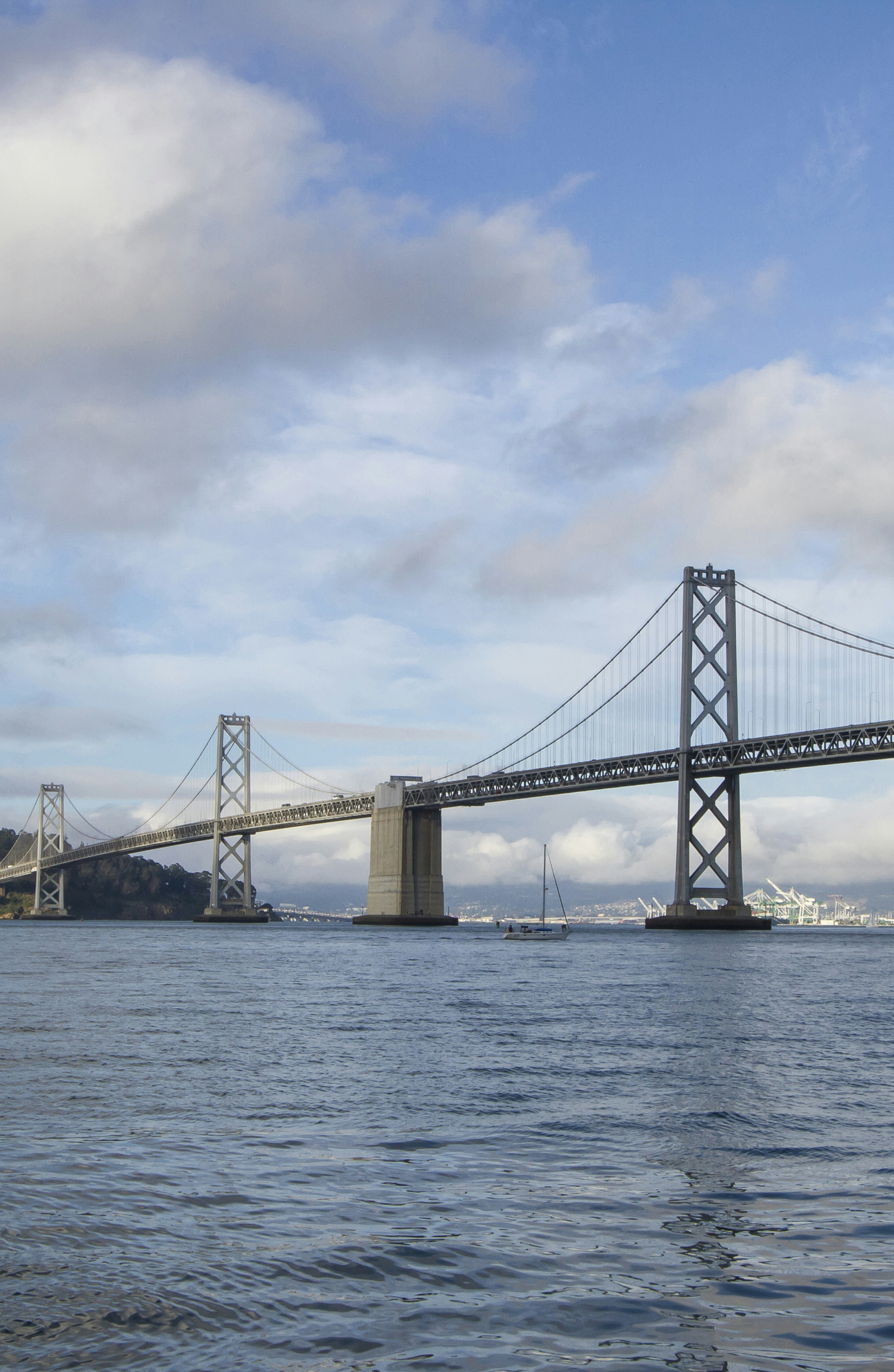 The Bay Bridge stretches across the water, showcasing its intricate design against a backdrop of clouds and blue sky. A small sailboat glides beneath the structure.