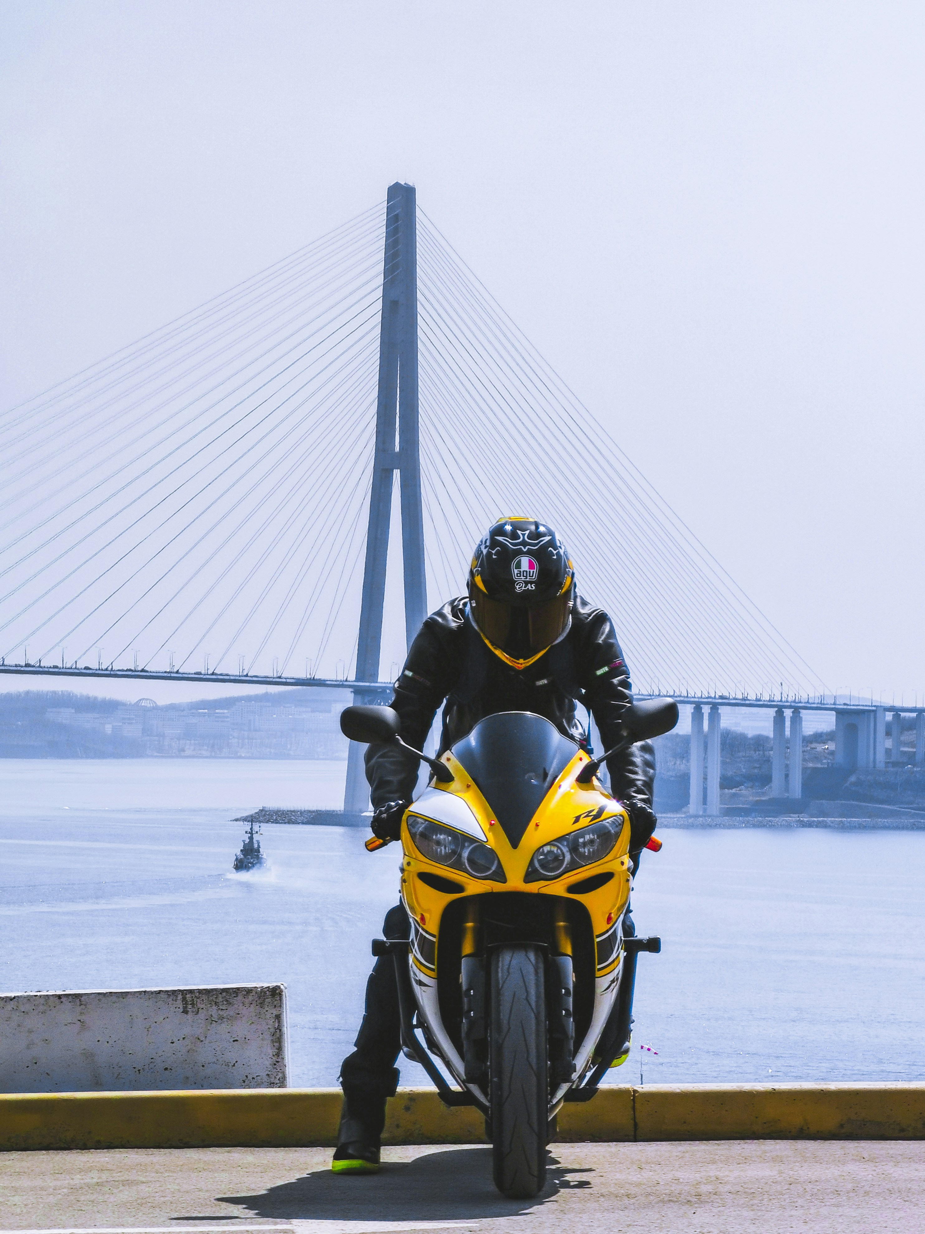 Rider in black gear on a yellow sportbike sits at a waterfront quay, with a cable-stayed bridge towering behind.