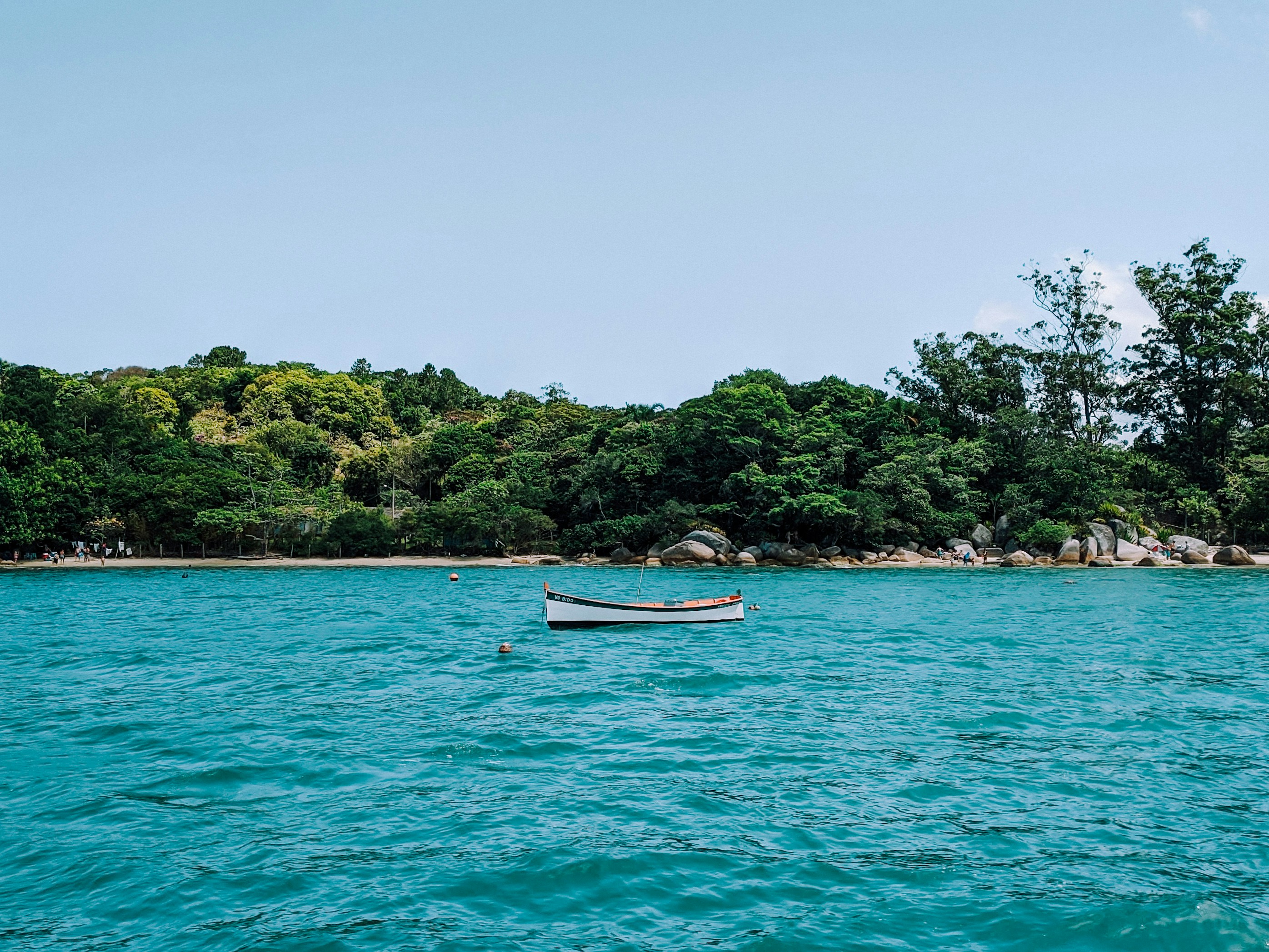 A lone boat floats on vibrant blue water against a backdrop of lush green shoreline under a clear sky.