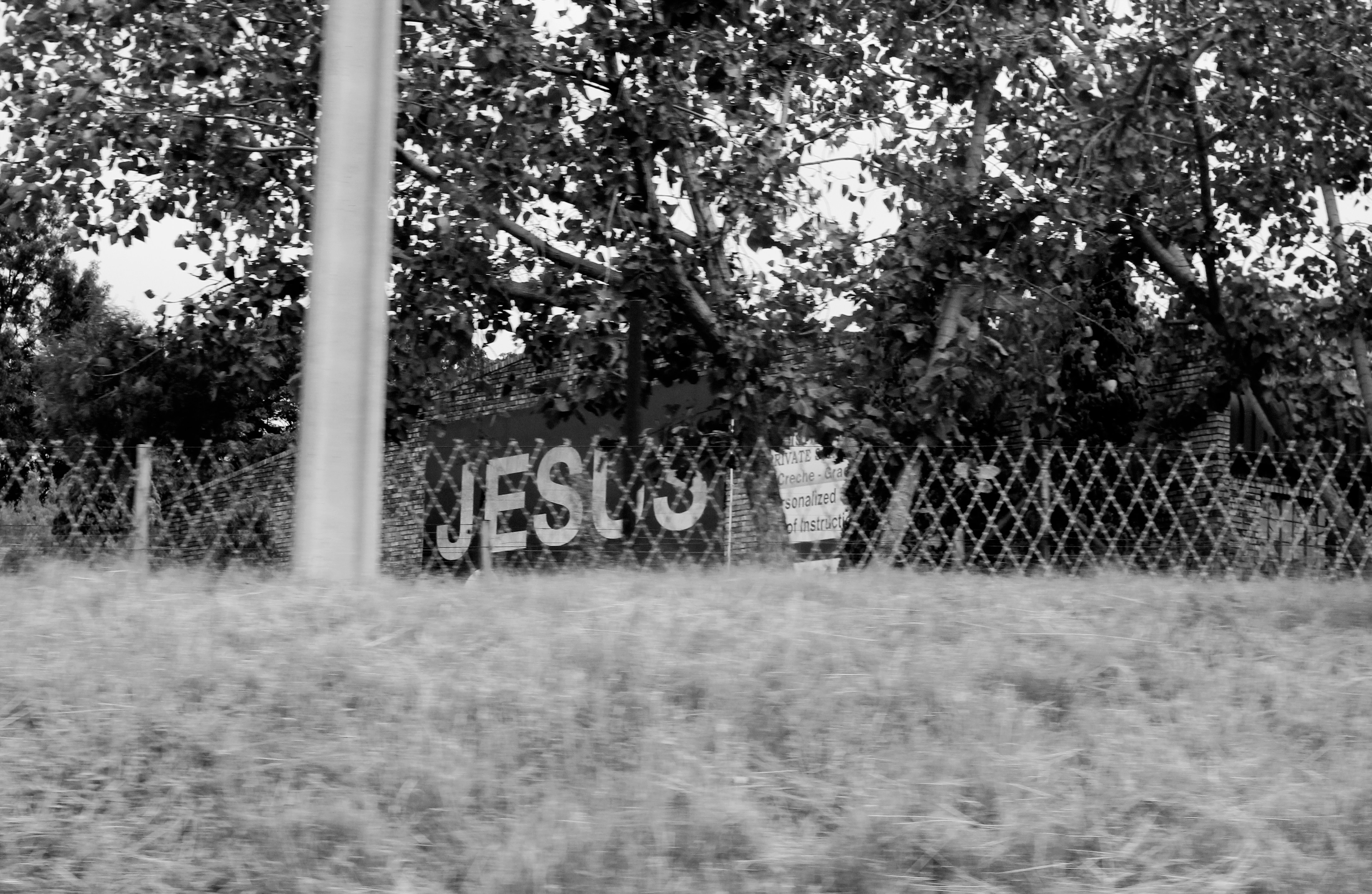 Grayscale chain link fence with 'JESUS' spelled out, set against a backdrop of trees.