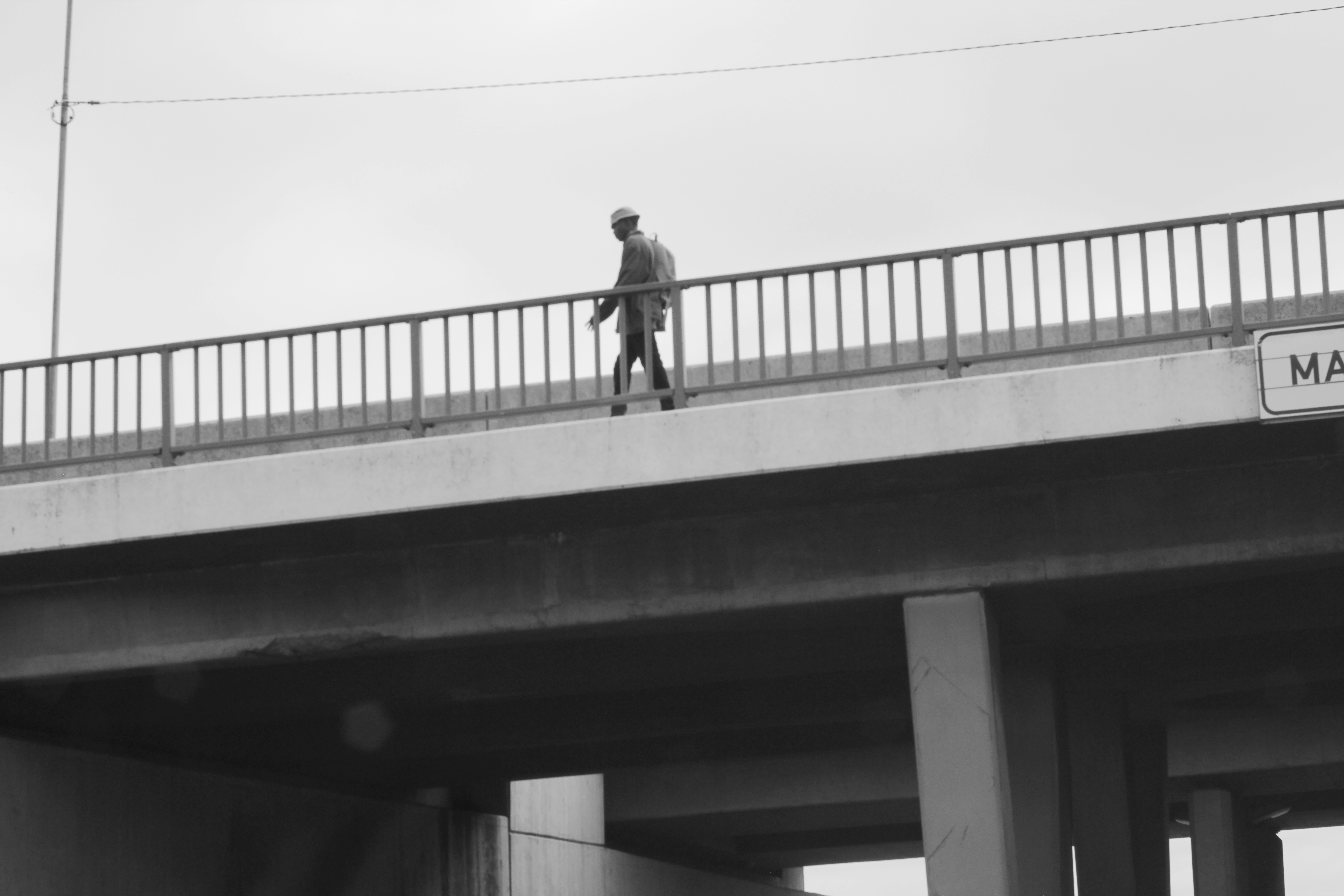 A lone figure walks along a concrete overpass, framed by the stark lines of urban architecture. The scene captures a moment of solitude in a bustling city environment.