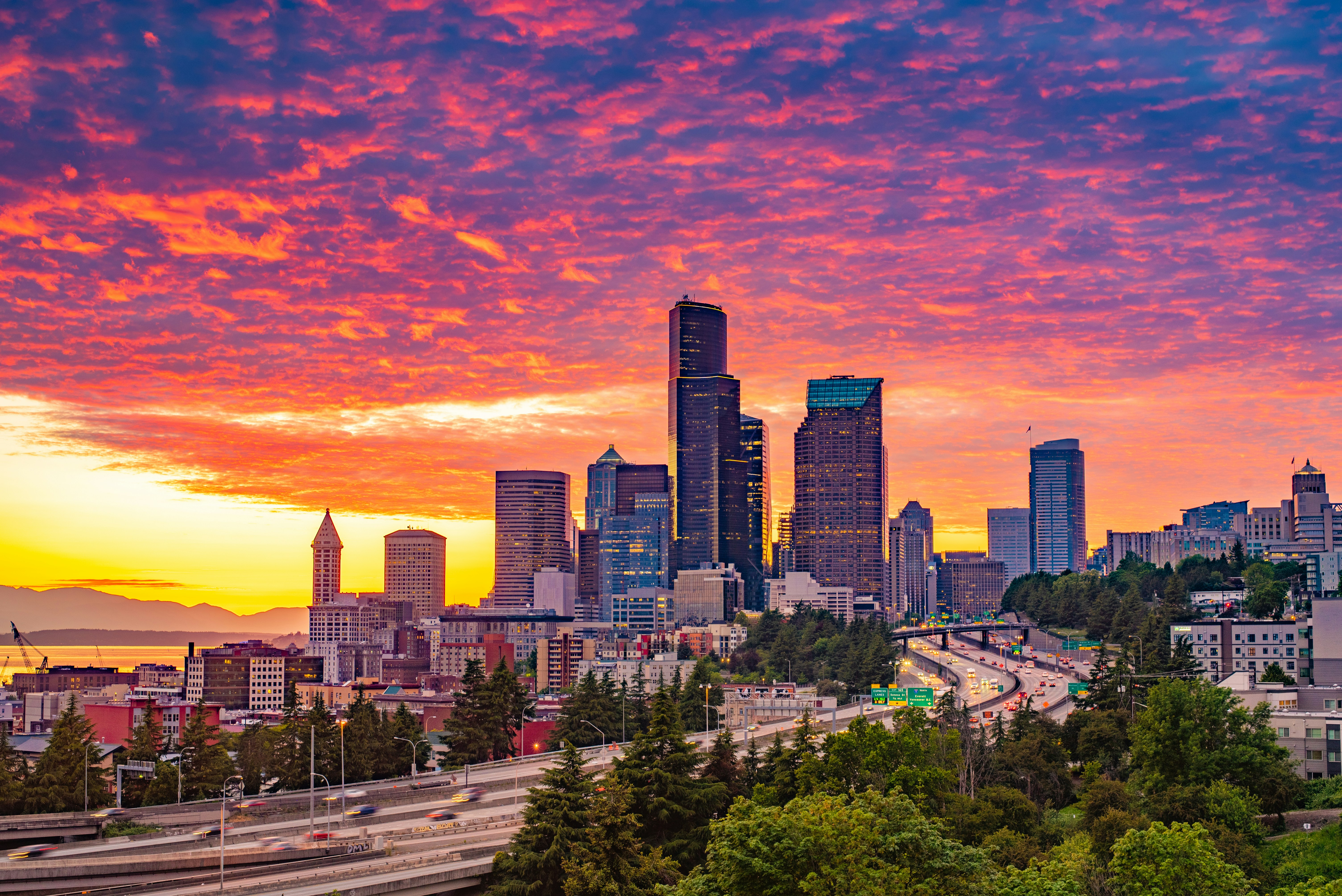 Vibrant sunset over a city skyline, showcasing towering skyscrapers and a winding highway framed by lush greenery.