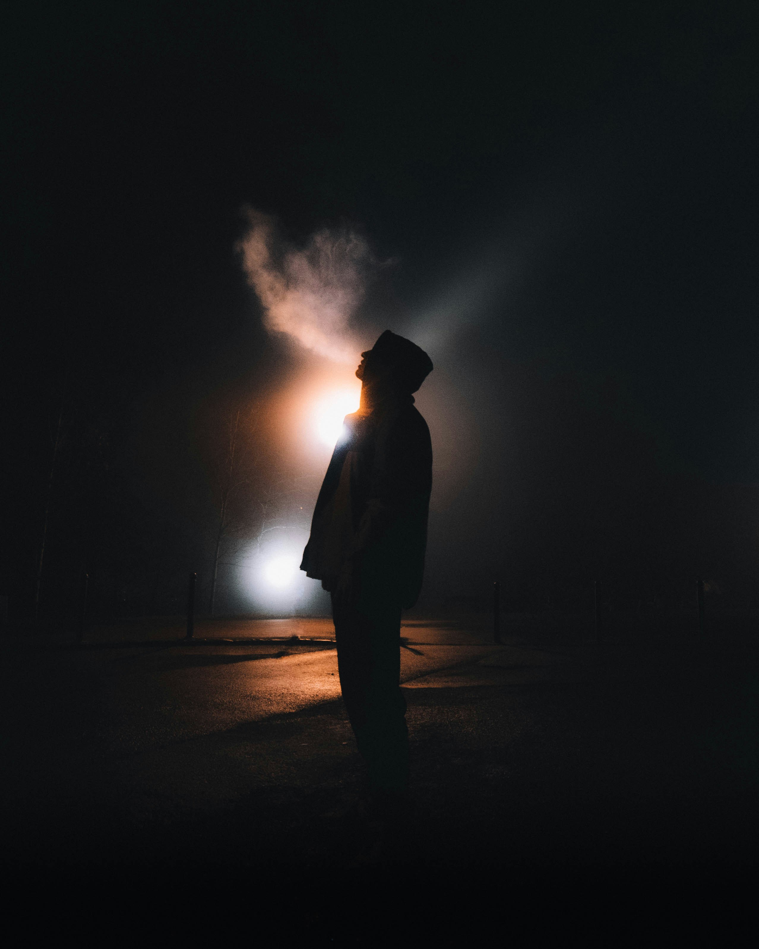 Silhouette of a person exhaling smoke, illuminated by distant lights in a foggy night setting.