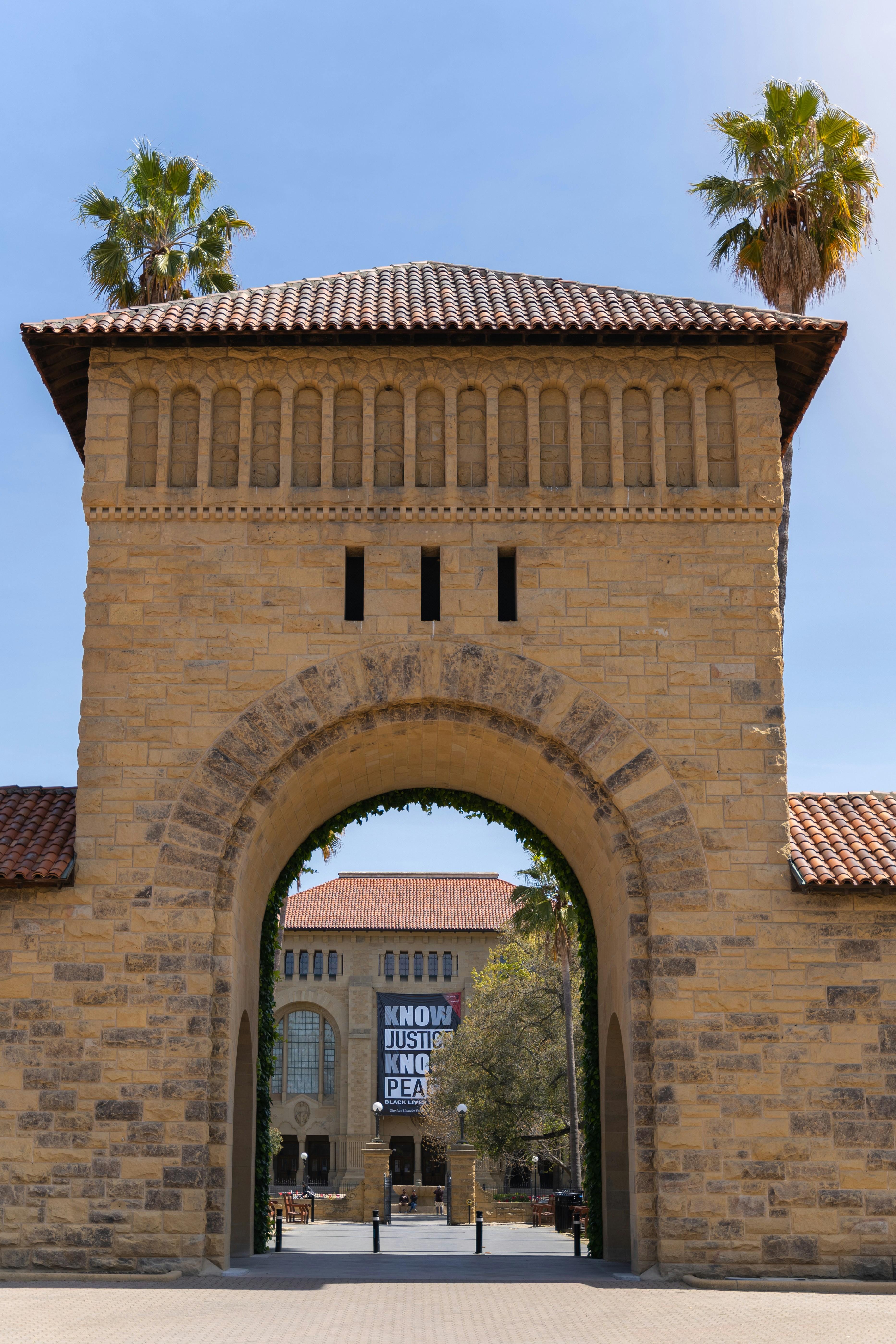 Historic stone archway framed by palm trees, leading to a university courtyard with a prominent banner. The architecture showcases classic design elements.