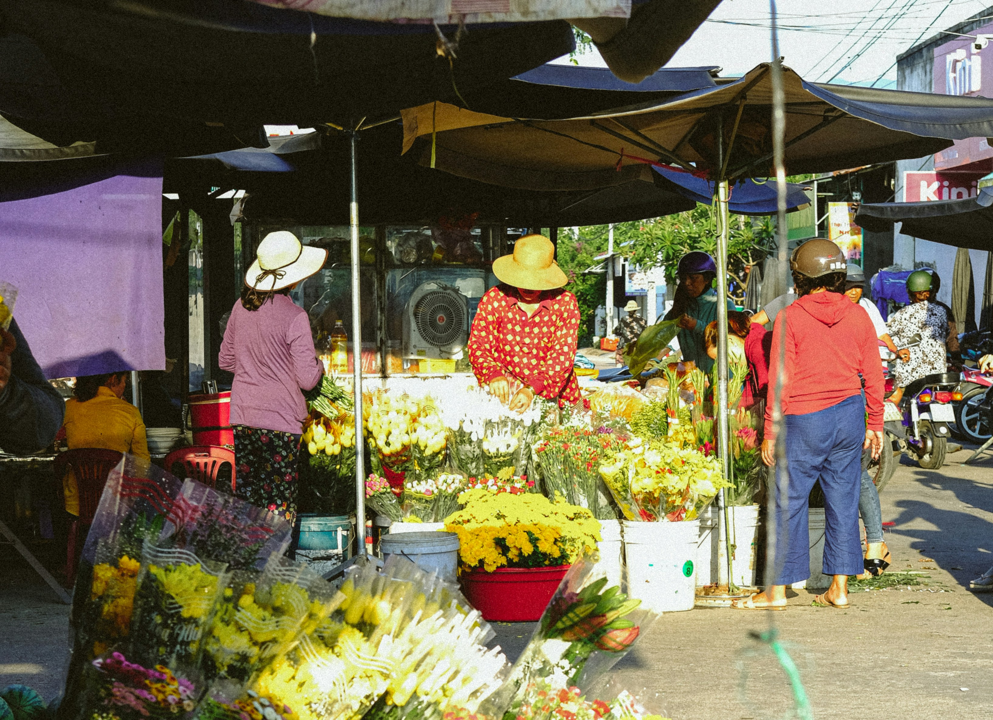 Flower vendors engage in lively trade under shaded stalls at a bustling market. Bright blooms create a colorful tapestry against the backdrop of daily life.