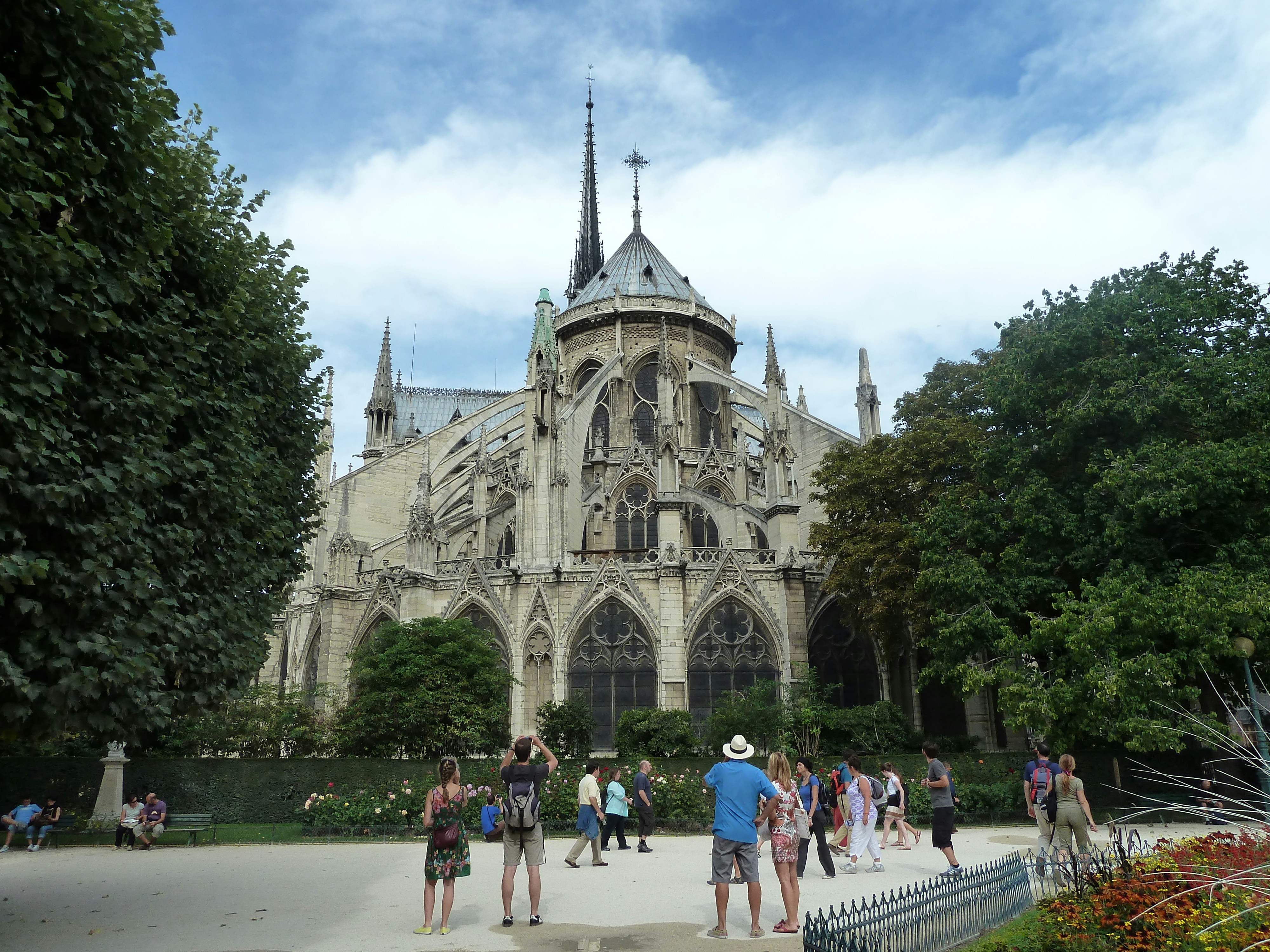 Gothic cathedral facade with rose window and spires dominates a sunlit plaza, framed by trees and visitors.