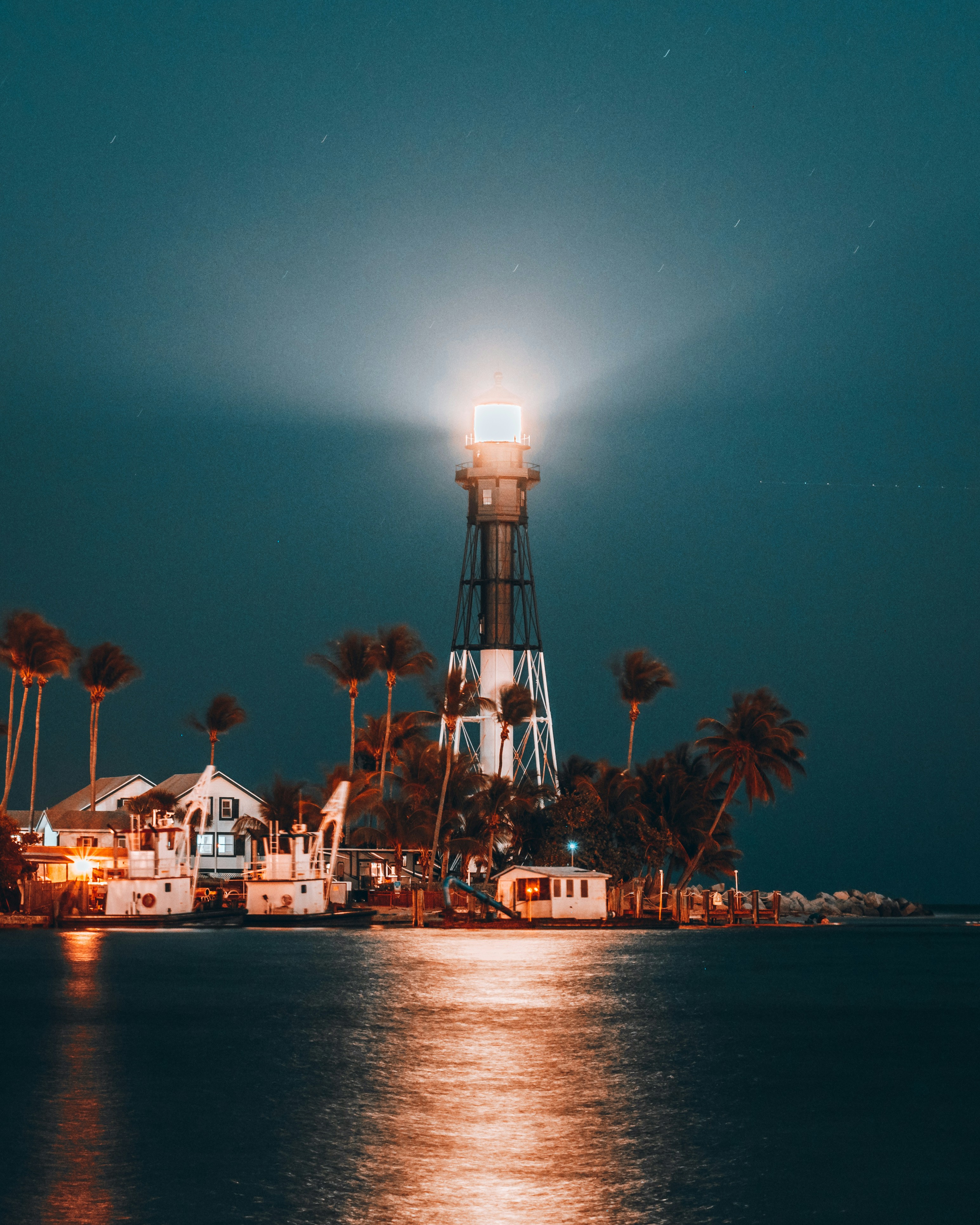 Lighthouse illuminating a tranquil coastal scene with palm trees under a night sky.