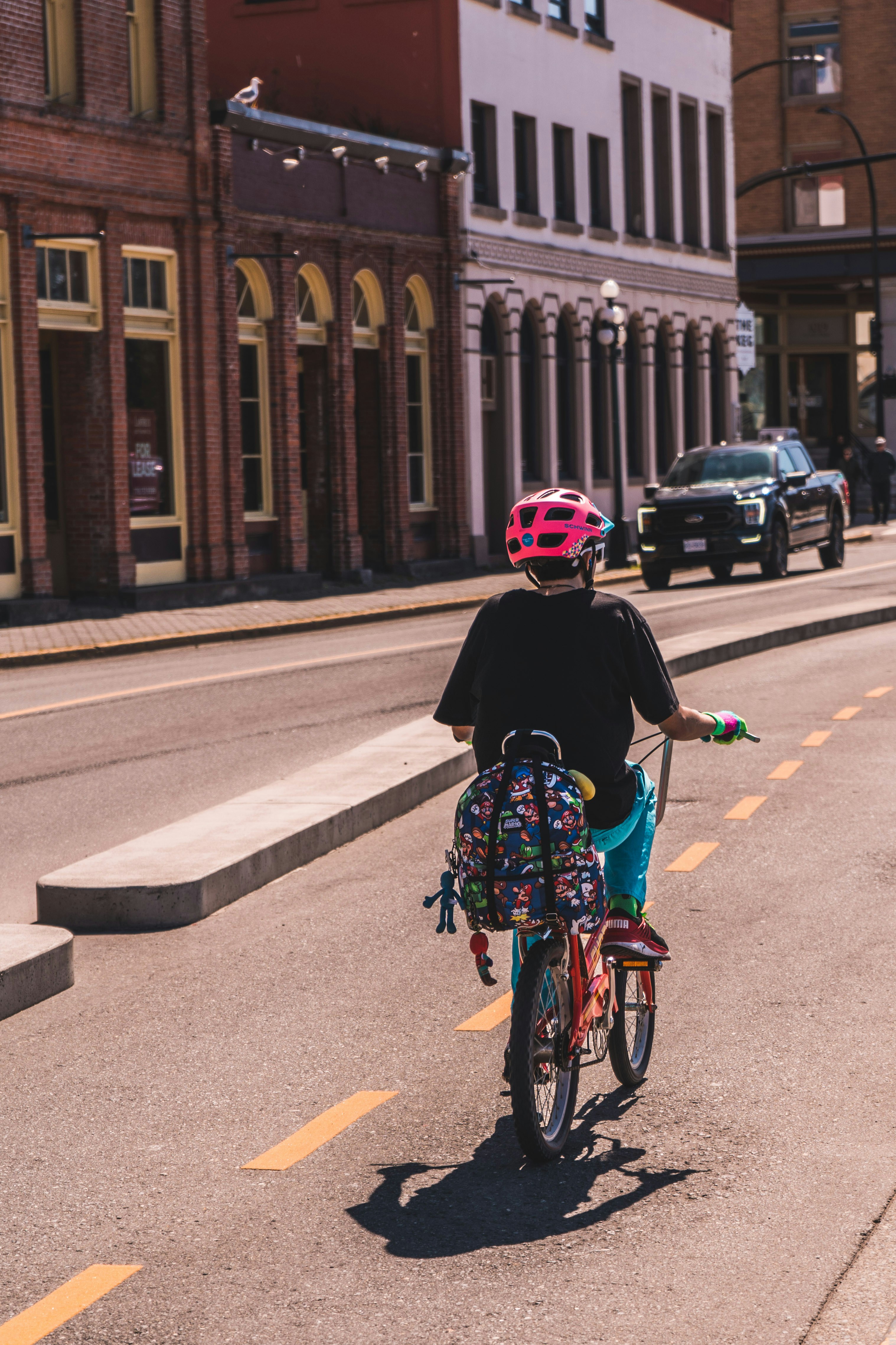 Child riding a bicycle along a city street, wearing a pink helmet and colorful backpack. Sunlight casts shadows on the pavement.