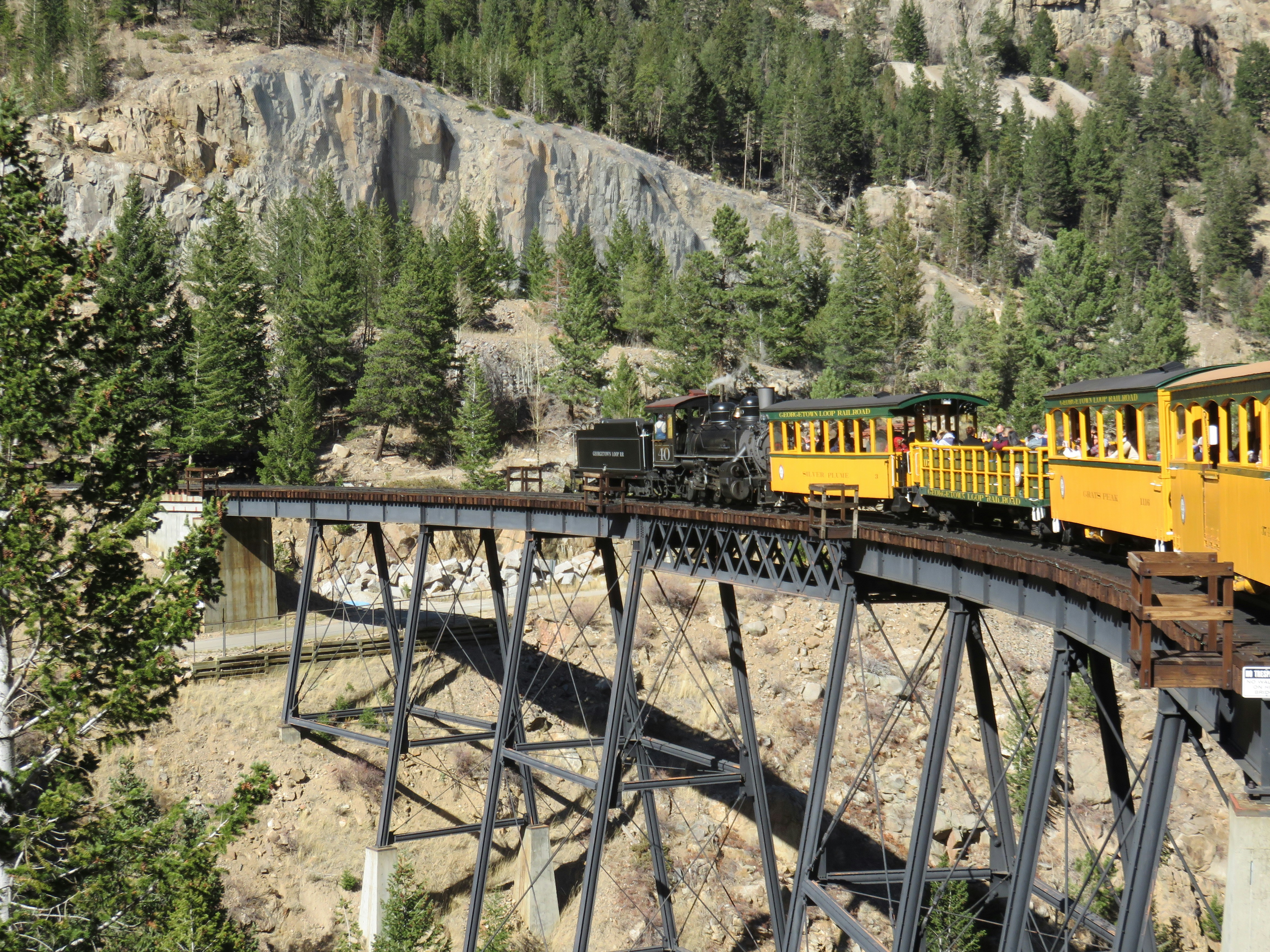 A vintage steam train crosses a trestle bridge surrounded by lush pine trees and rocky cliffs.
