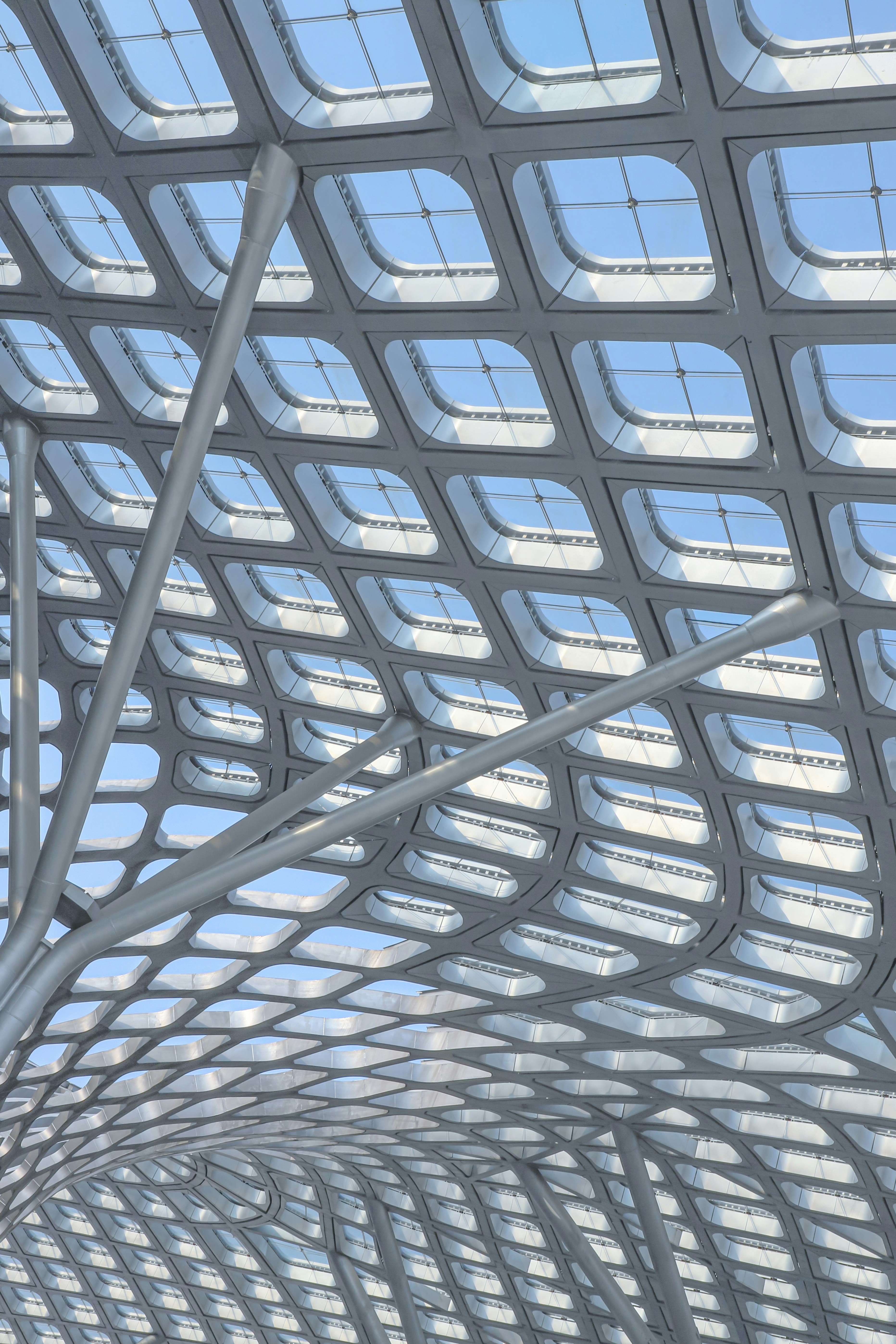 Intricate latticework of a modern architectural ceiling, showcasing a blend of steel and glass against a clear blue sky.