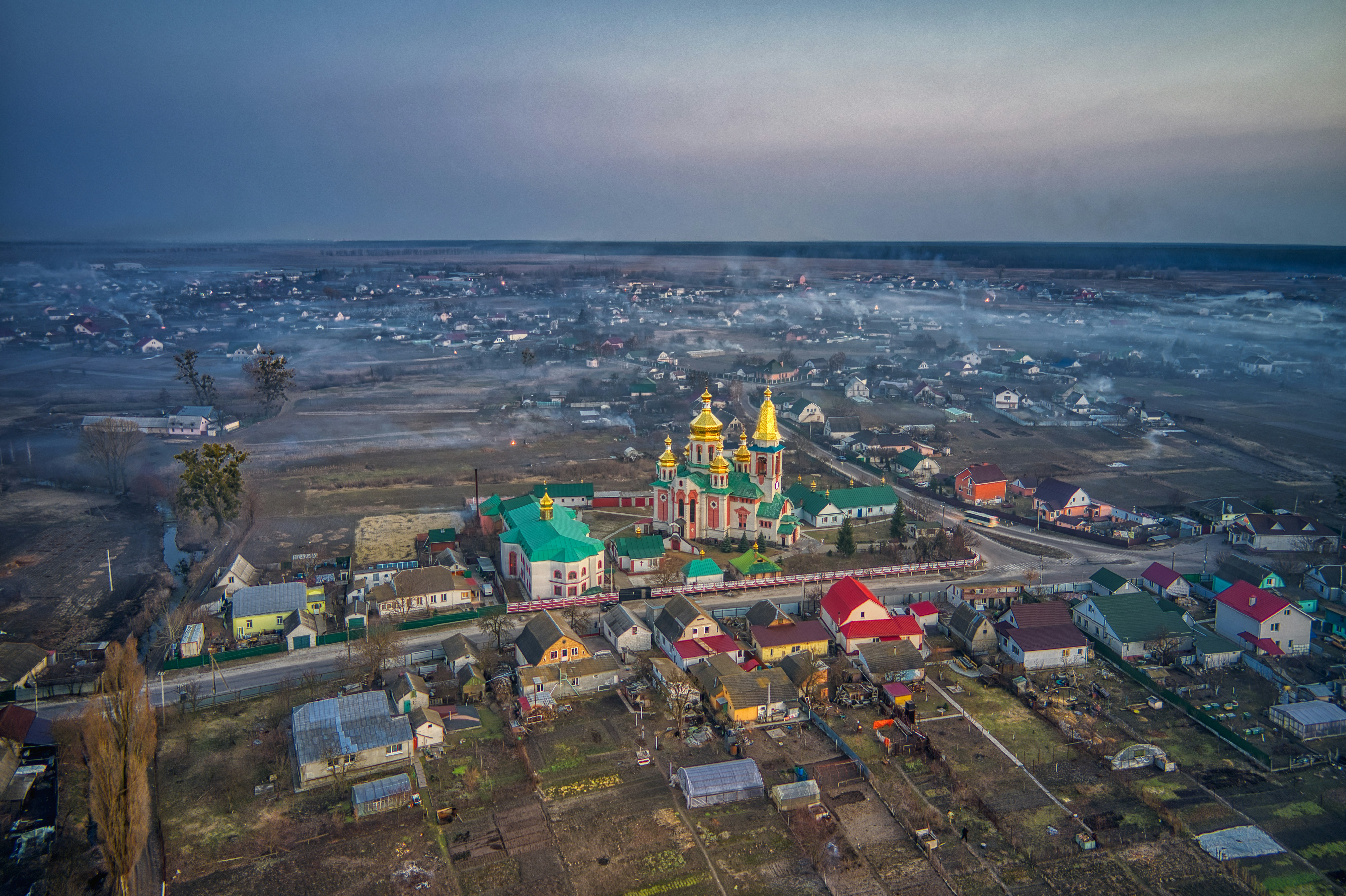 Aerial view of a colorful church with golden domes surrounded by a rural landscape of houses and fields, shrouded in mist.