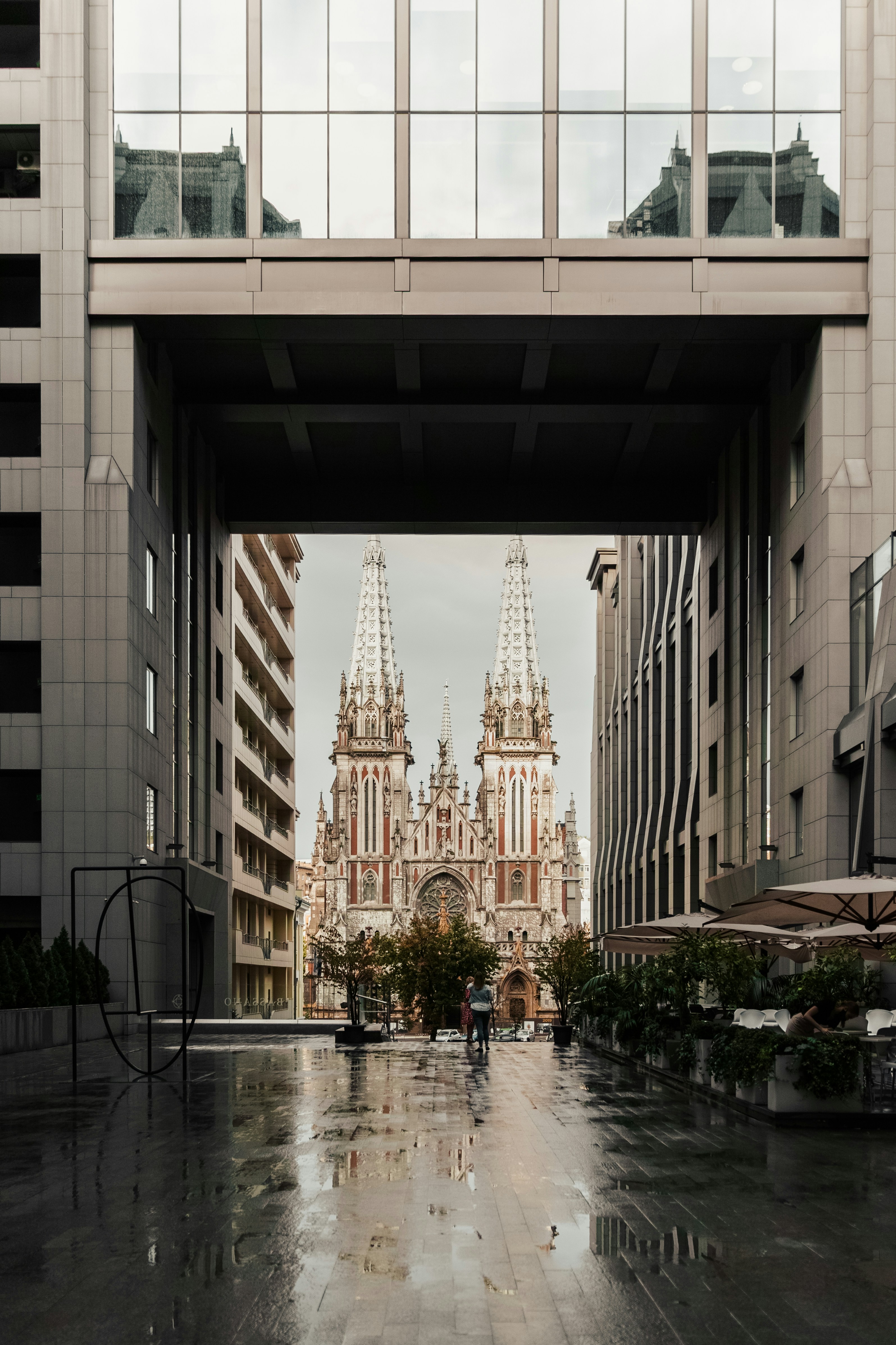 A narrow urban passage framed by modern buildings opens toward a Gothic cathedral with twin spires. Wet pavement reflects the surrounding architecture as pedestrians traverse the space.