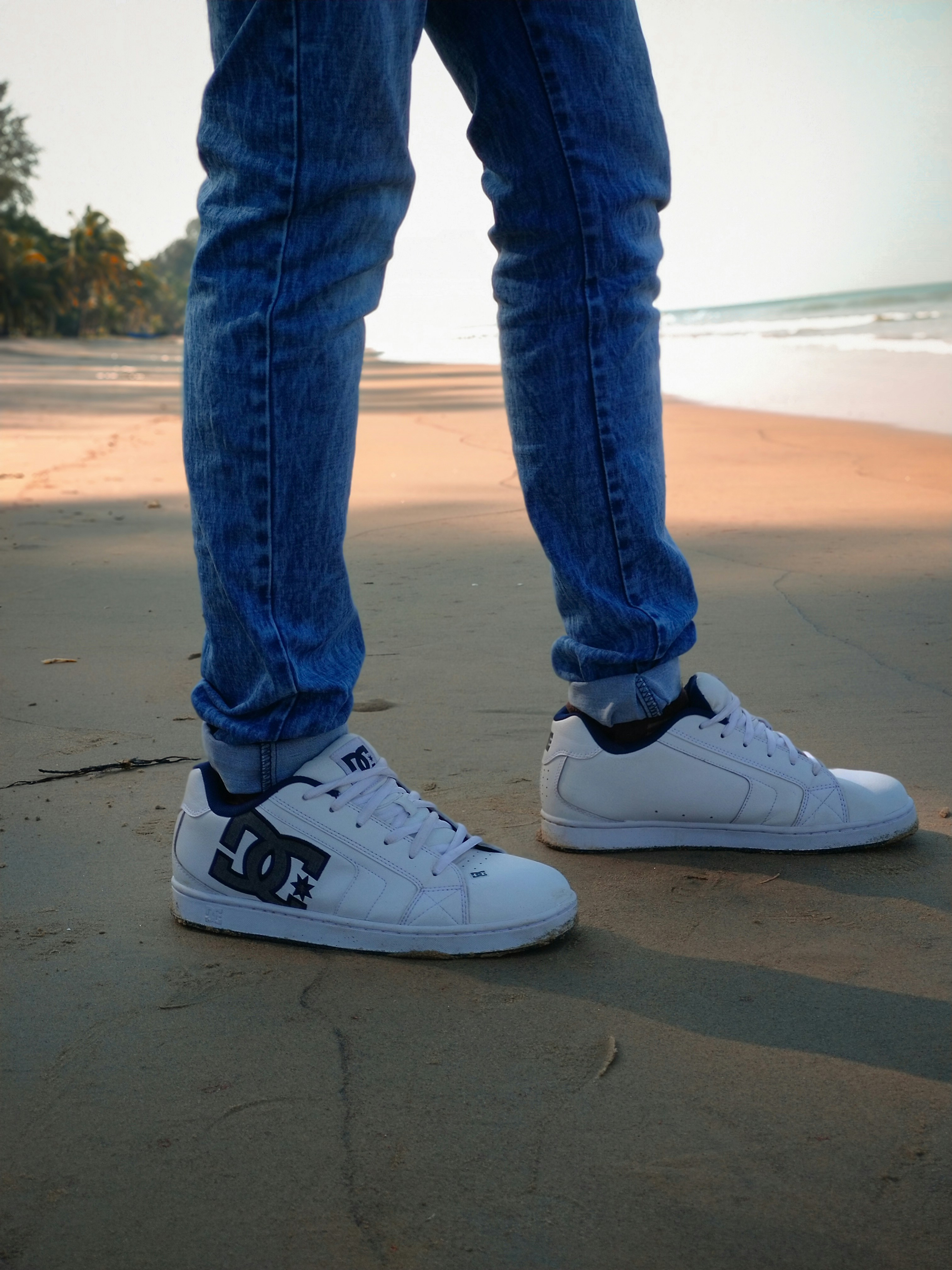 White sneakers with a logo stand on a sandy beach, with gentle waves in the background and lush greenery lining the shore.