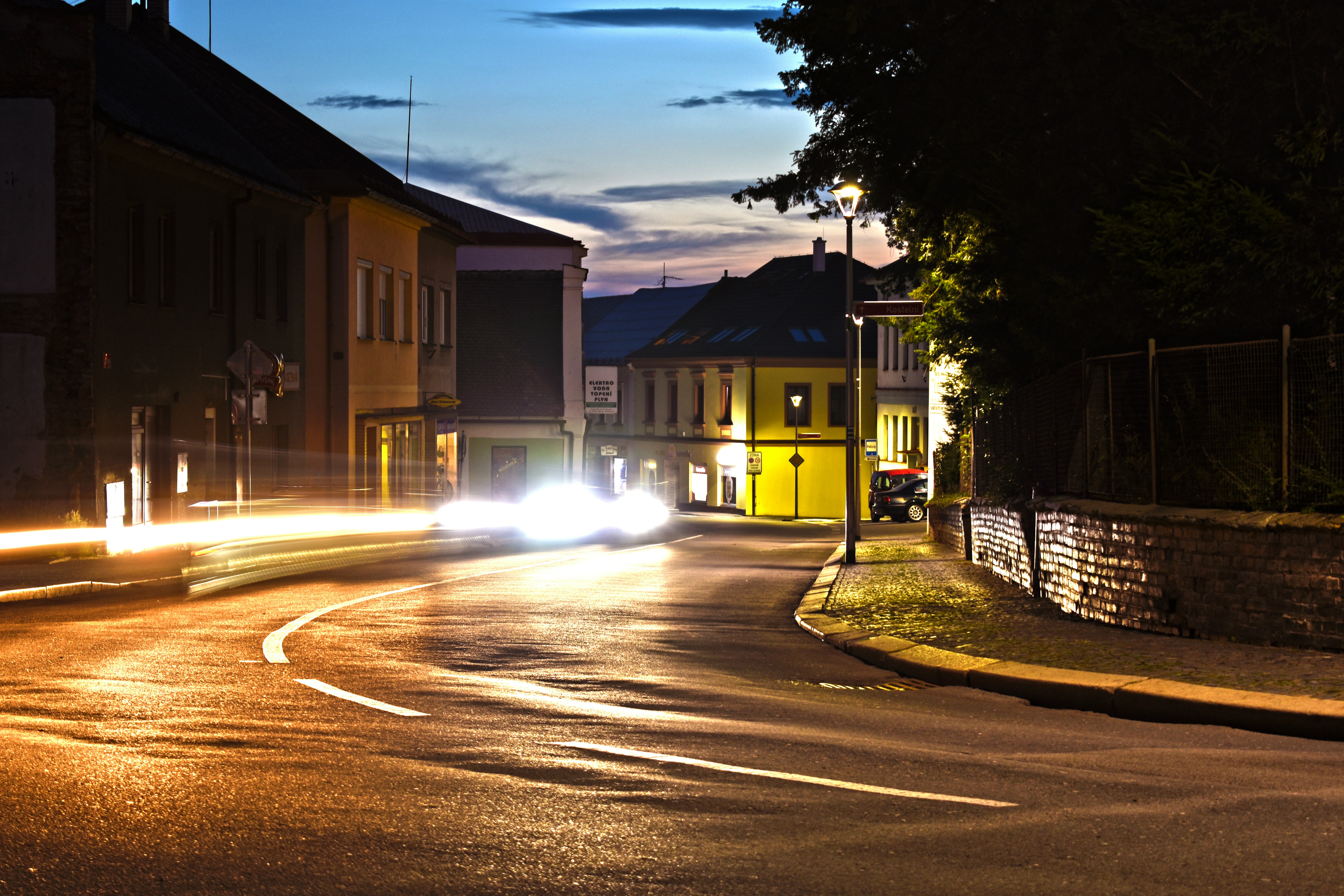 Curving street illuminated by vehicle headlights, surrounded by quaint buildings under a twilight sky.