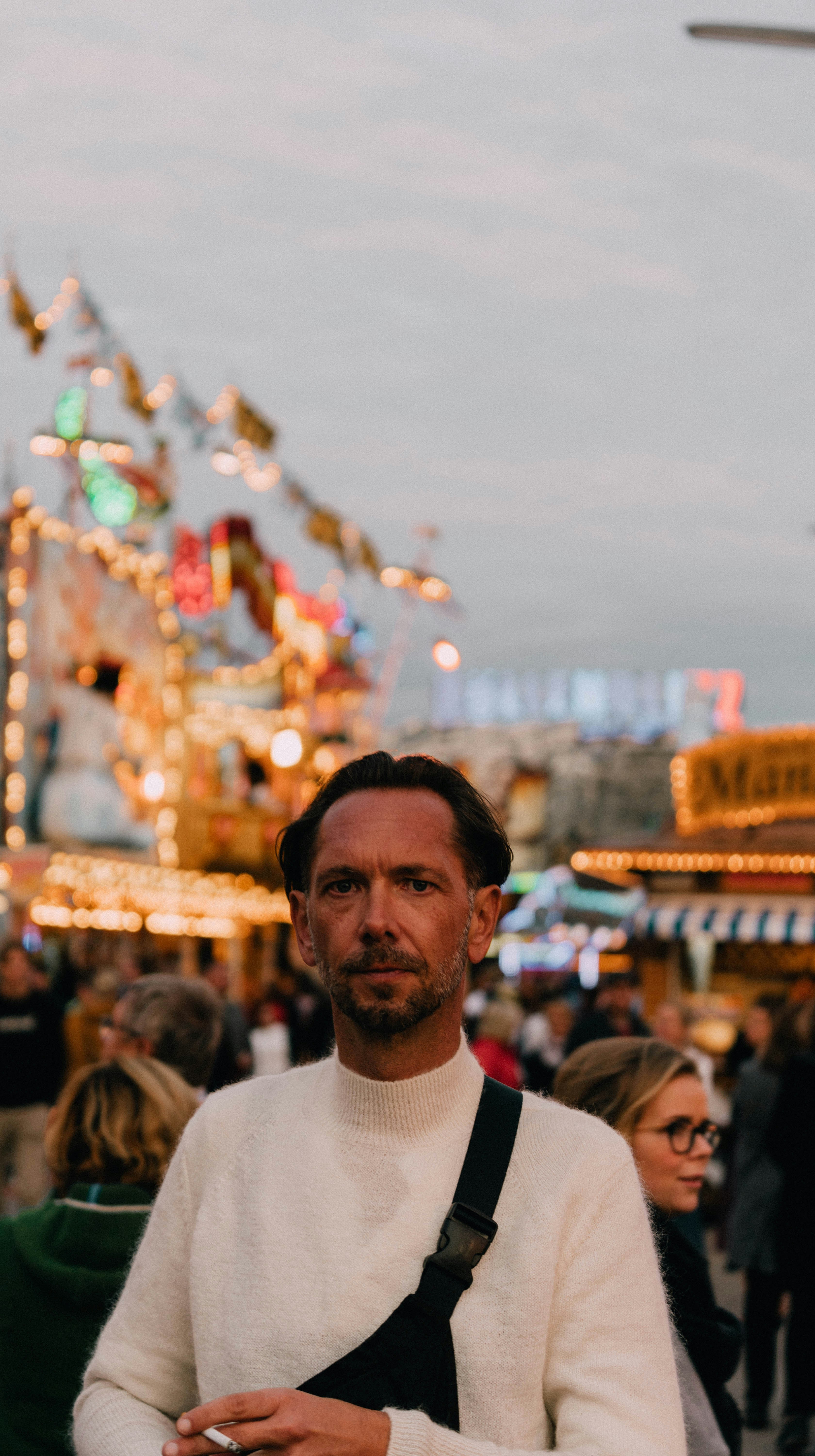 Man in a white sweater stands confidently among a bustling fairground illuminated by colorful lights.