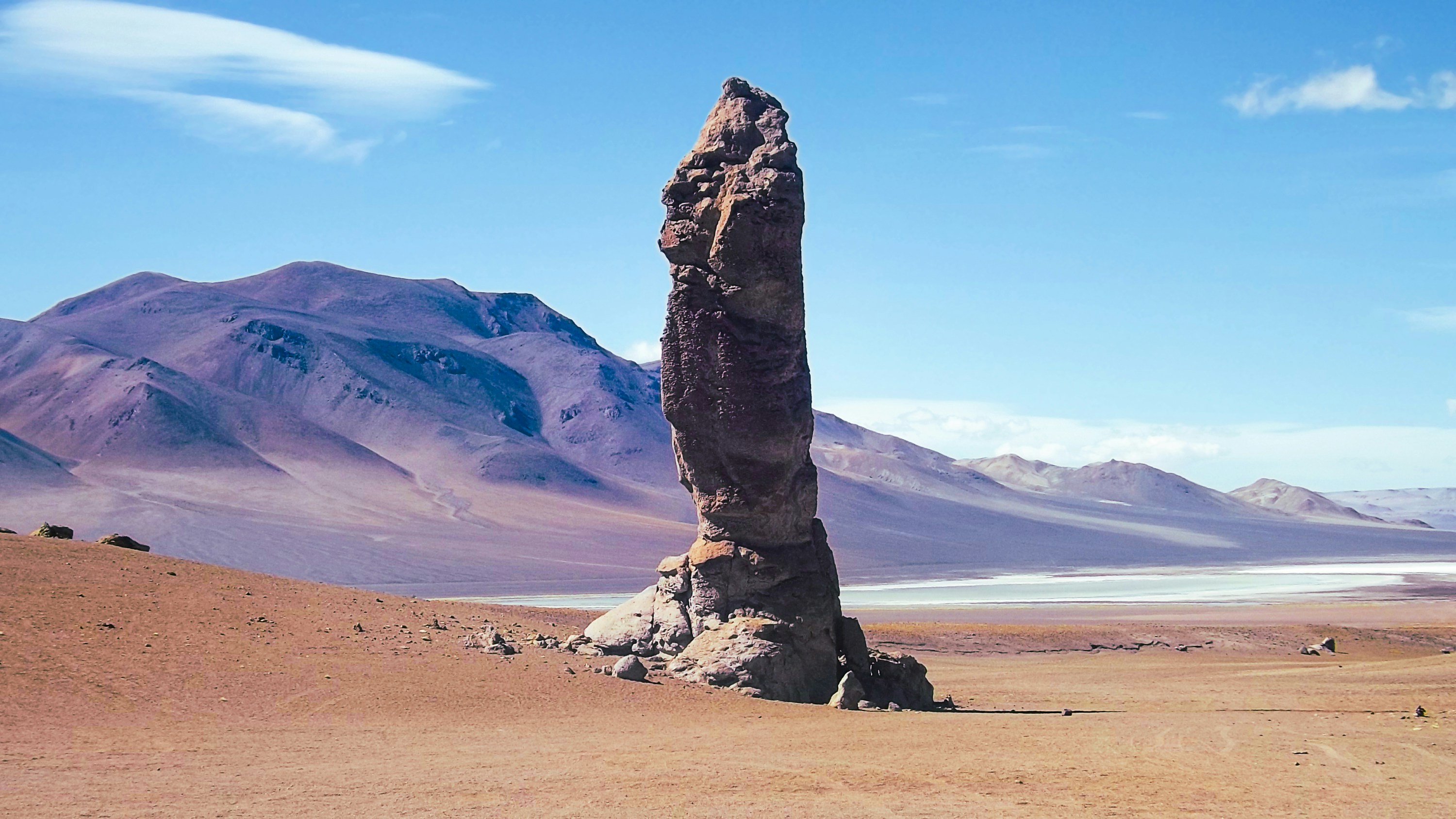 A solitary rock pillar rises from a barren desert plain with distant purple mountains under a clear blue sky. The composition emphasizes stark geology and vast emptiness.