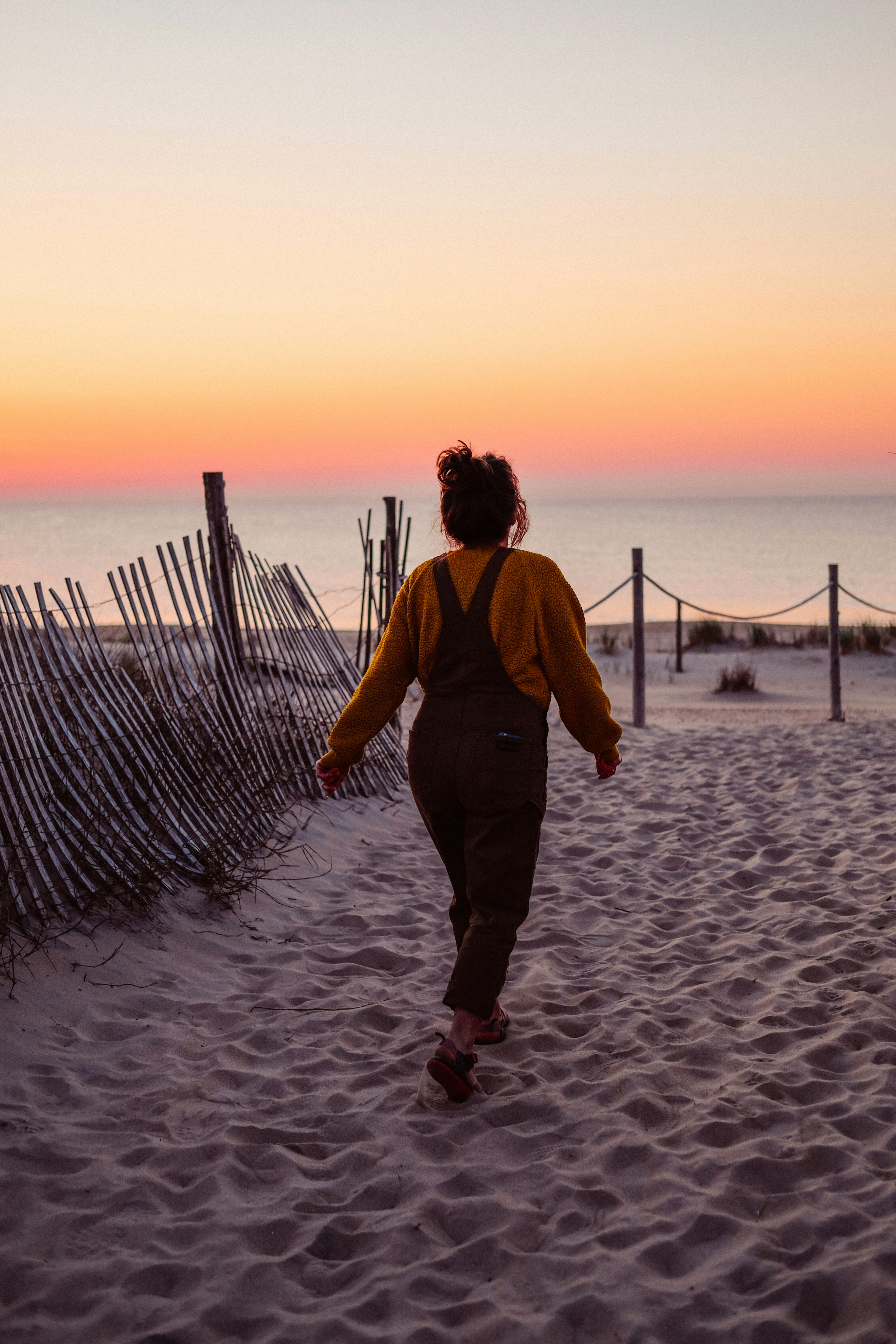 A person walks along a sandy beach path, framed by weathered fencing, as the sun sets over the tranquil ocean waters.