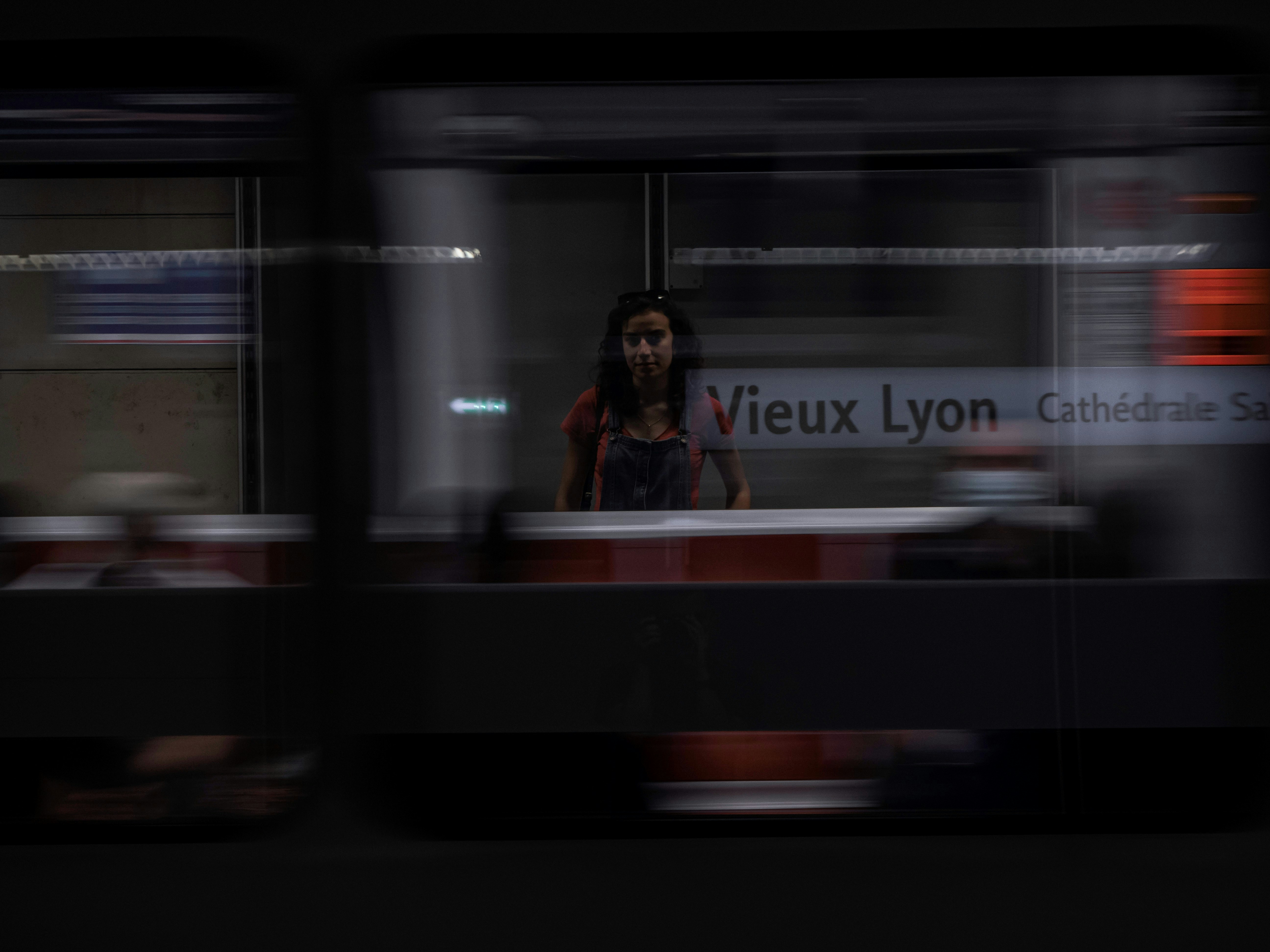 A solitary figure stands in a subway station, framed by the motion of passing trains, highlighting the contrast between stillness and speed.