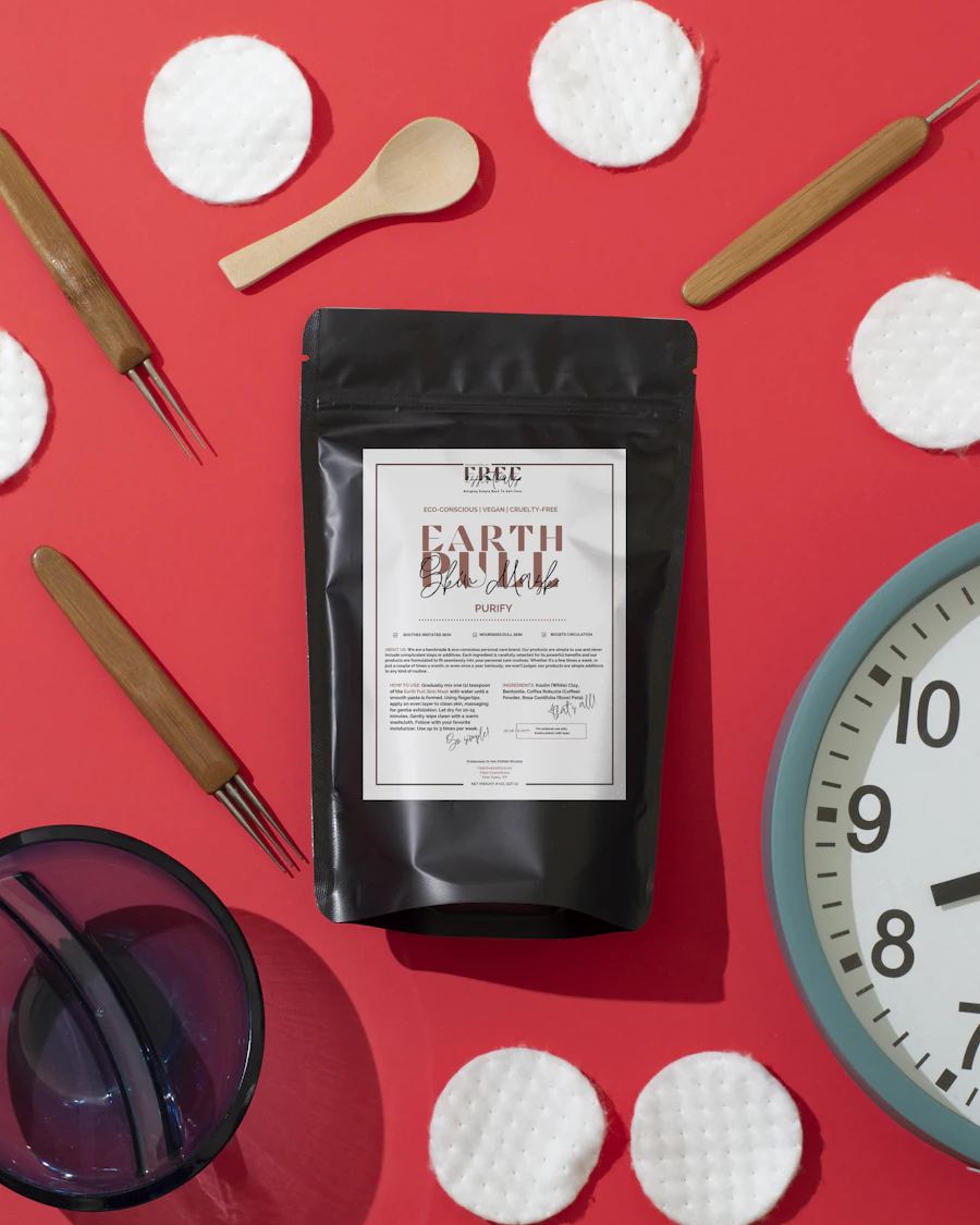Black resealable skincare pouch centered on a bright red background, surrounded by cotton pads, wooden tools, a small bowl, and the edge of a clock. The arrangement feels like a clean beauty product flat lay.