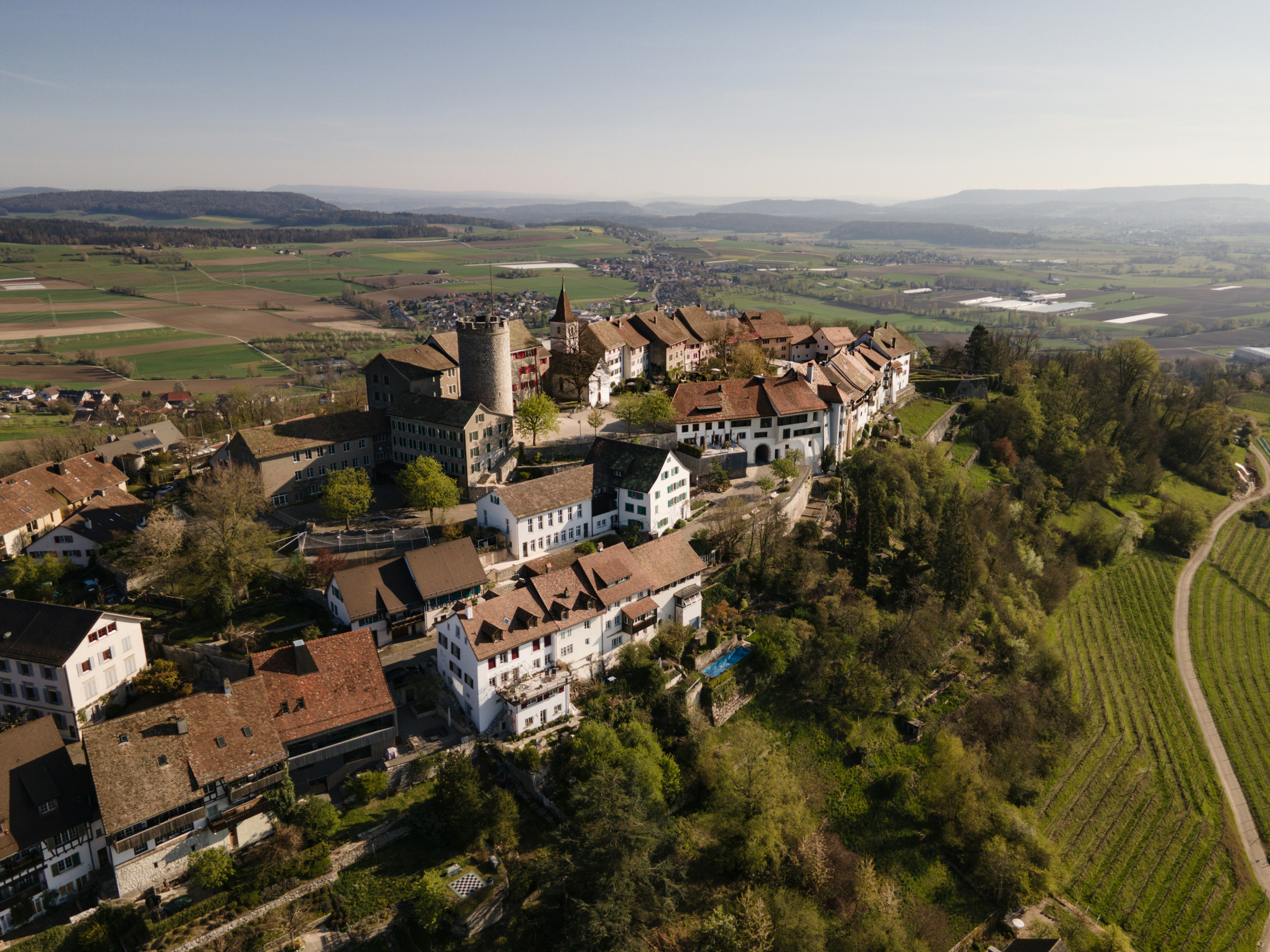 Aerial view of a quaint village nestled among rolling hills and vineyards, showcasing a blend of historical architecture and lush greenery.