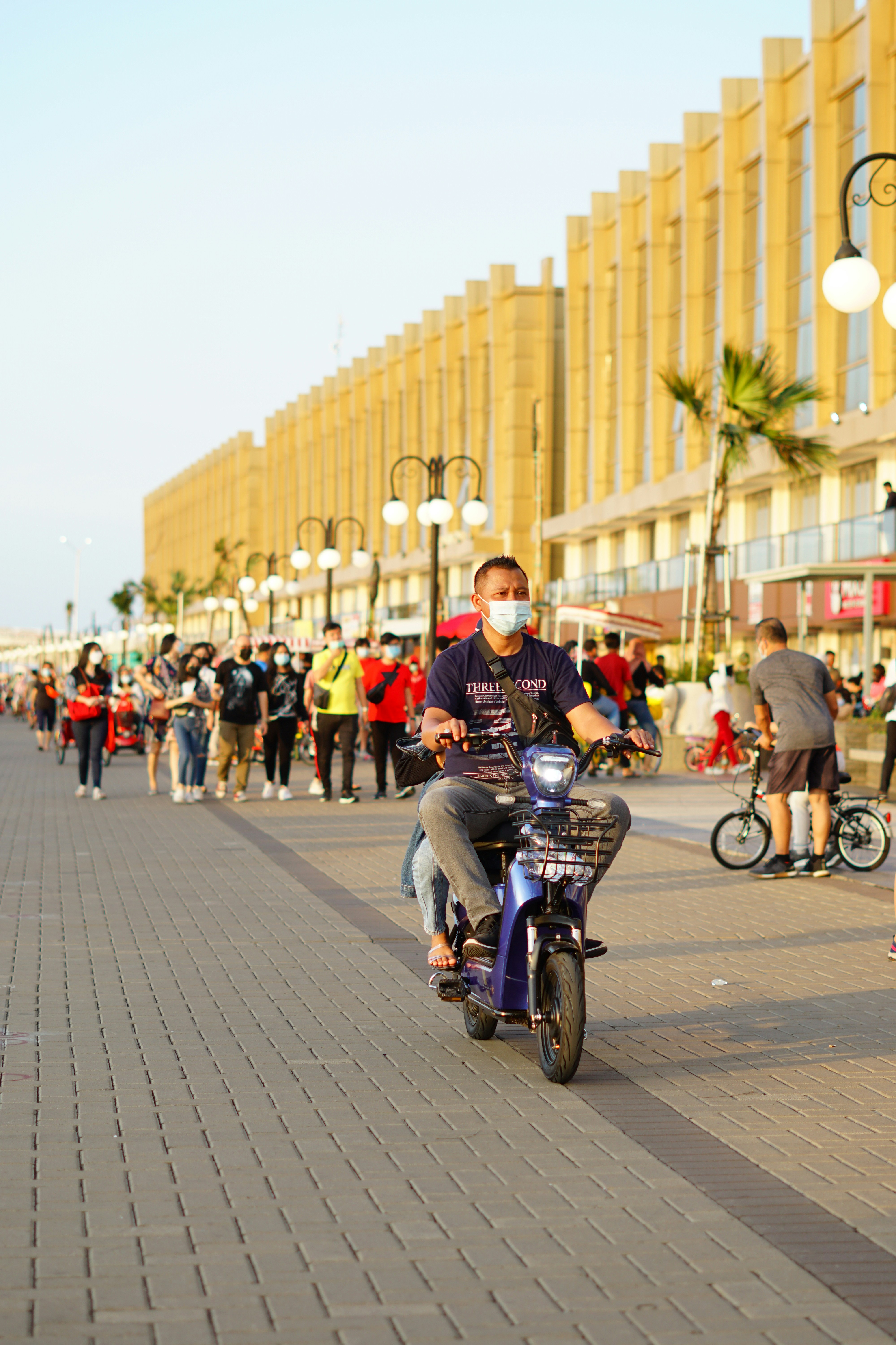 A man on an electric scooter navigates a busy promenade lined with pedestrians and palm trees during golden hour.