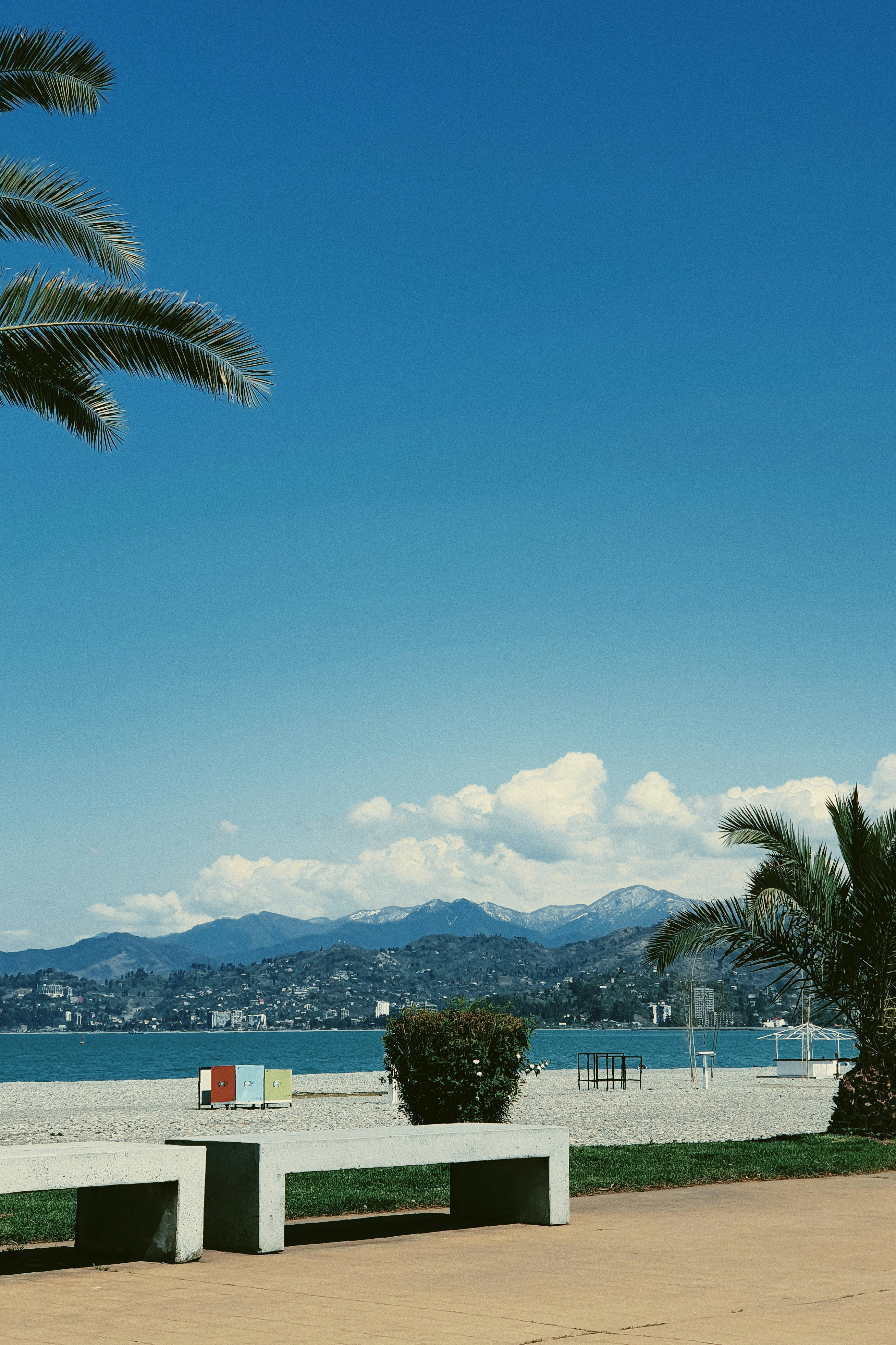 Concrete bench overlooking a tranquil beach scene with distant mountains and palm trees under a clear blue sky.