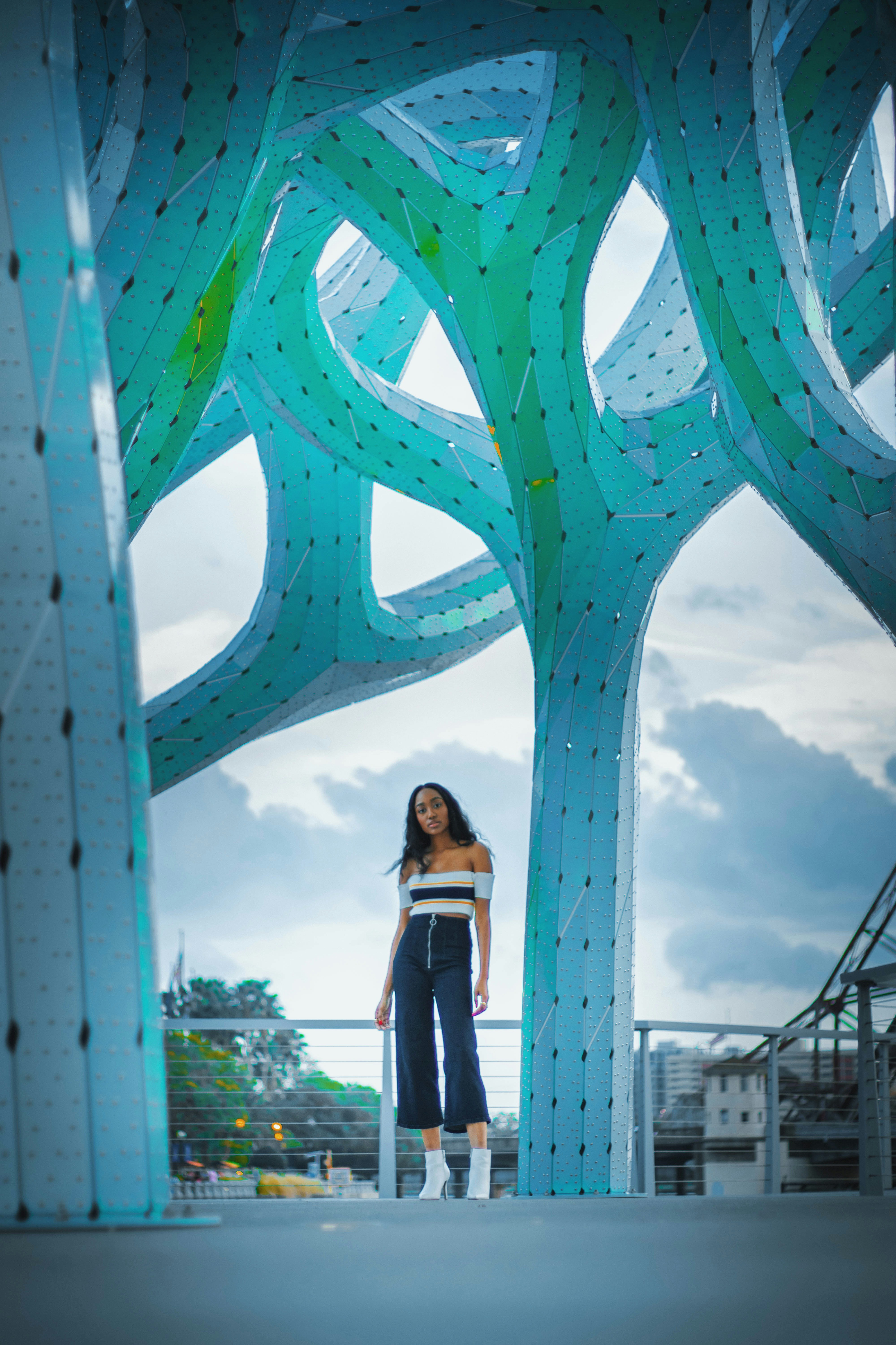 A woman stands gracefully beneath an intricate, blue-hued architectural installation, framed by swirling patterns against a cloudy sky.