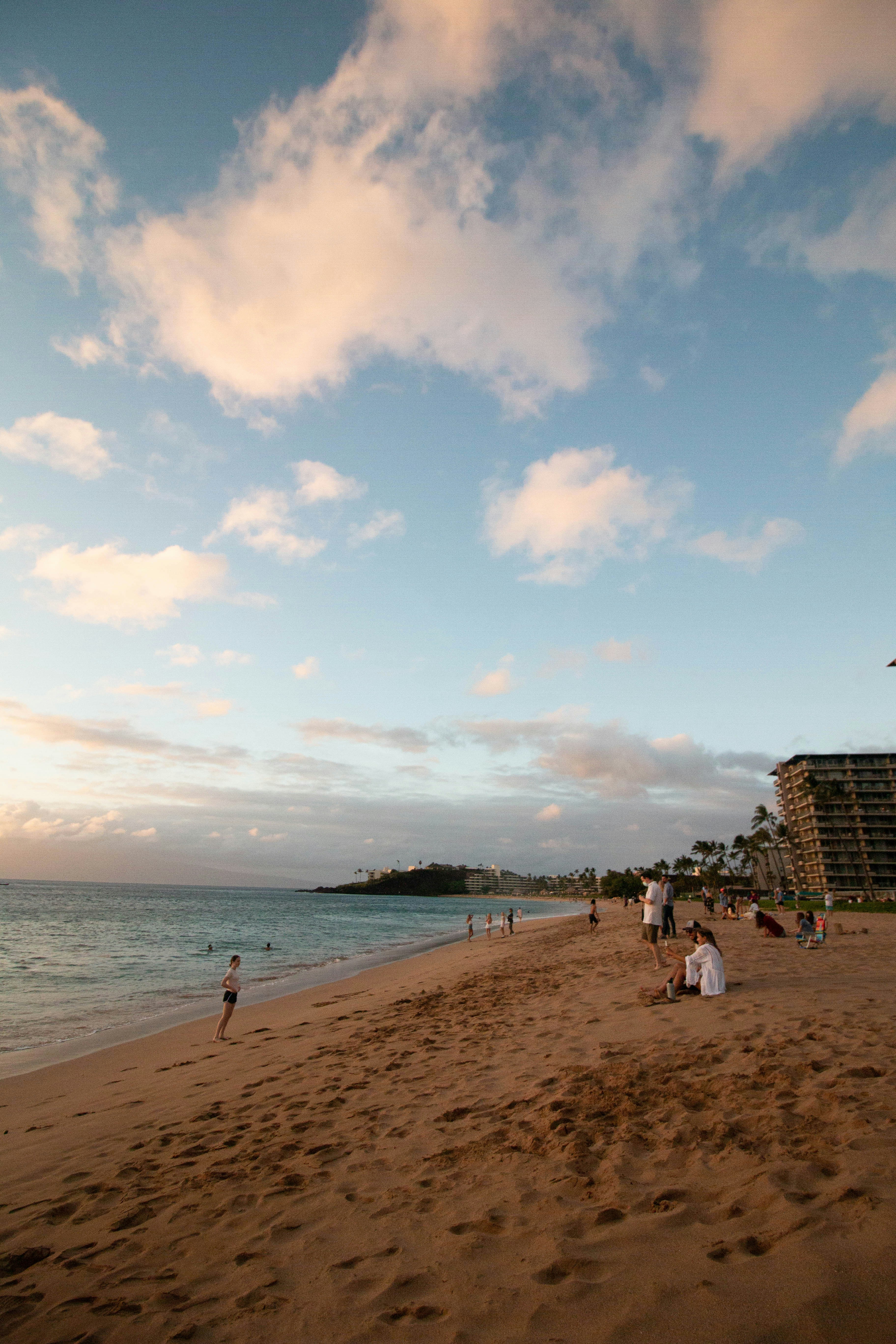 Golden sand beach at dusk with people enjoying the tranquil ocean waves and soft clouds above.