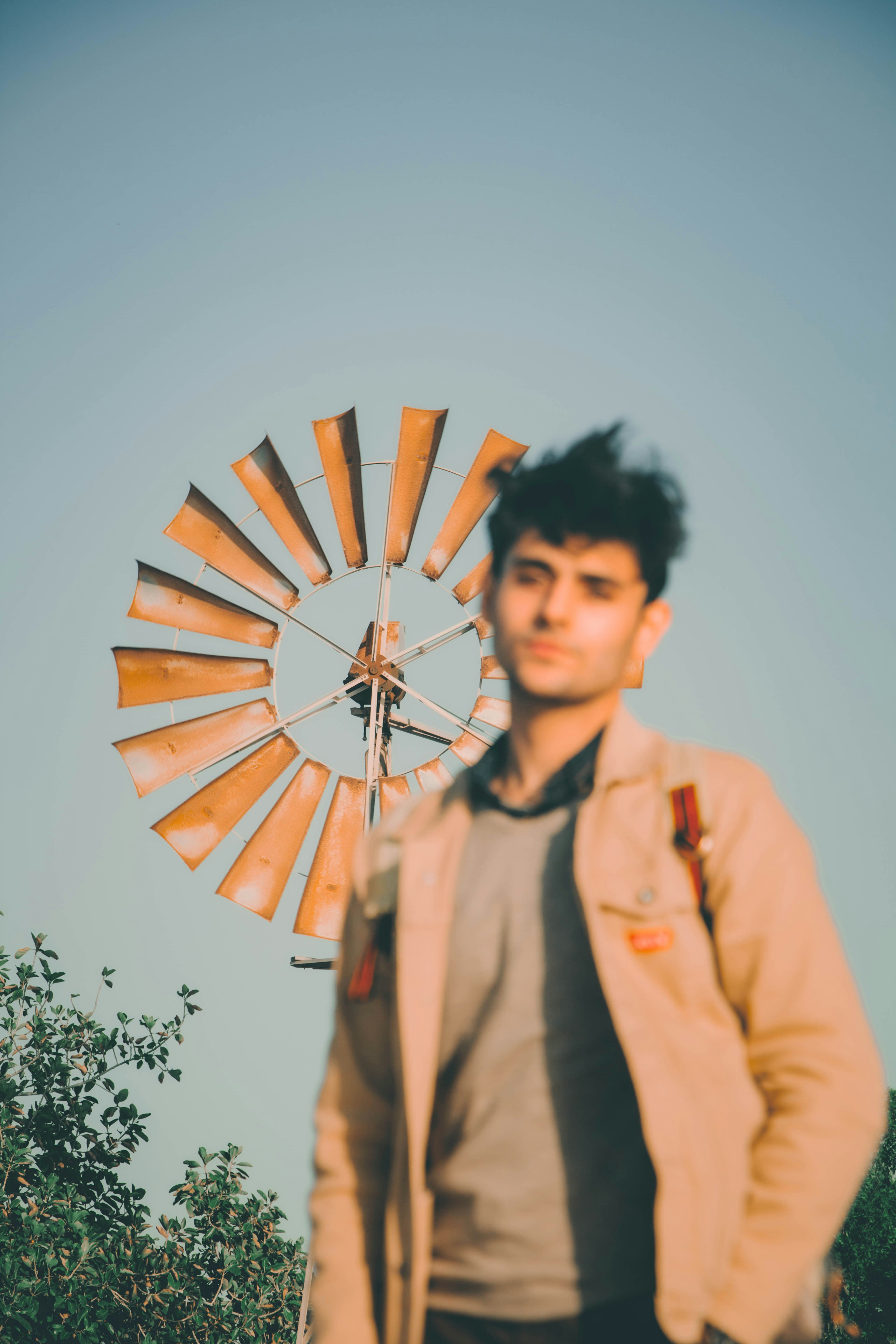 A young man stands blurred in the foreground, with a vintage windmill rotating in the background. The scene captures a serene moment in a rural landscape.