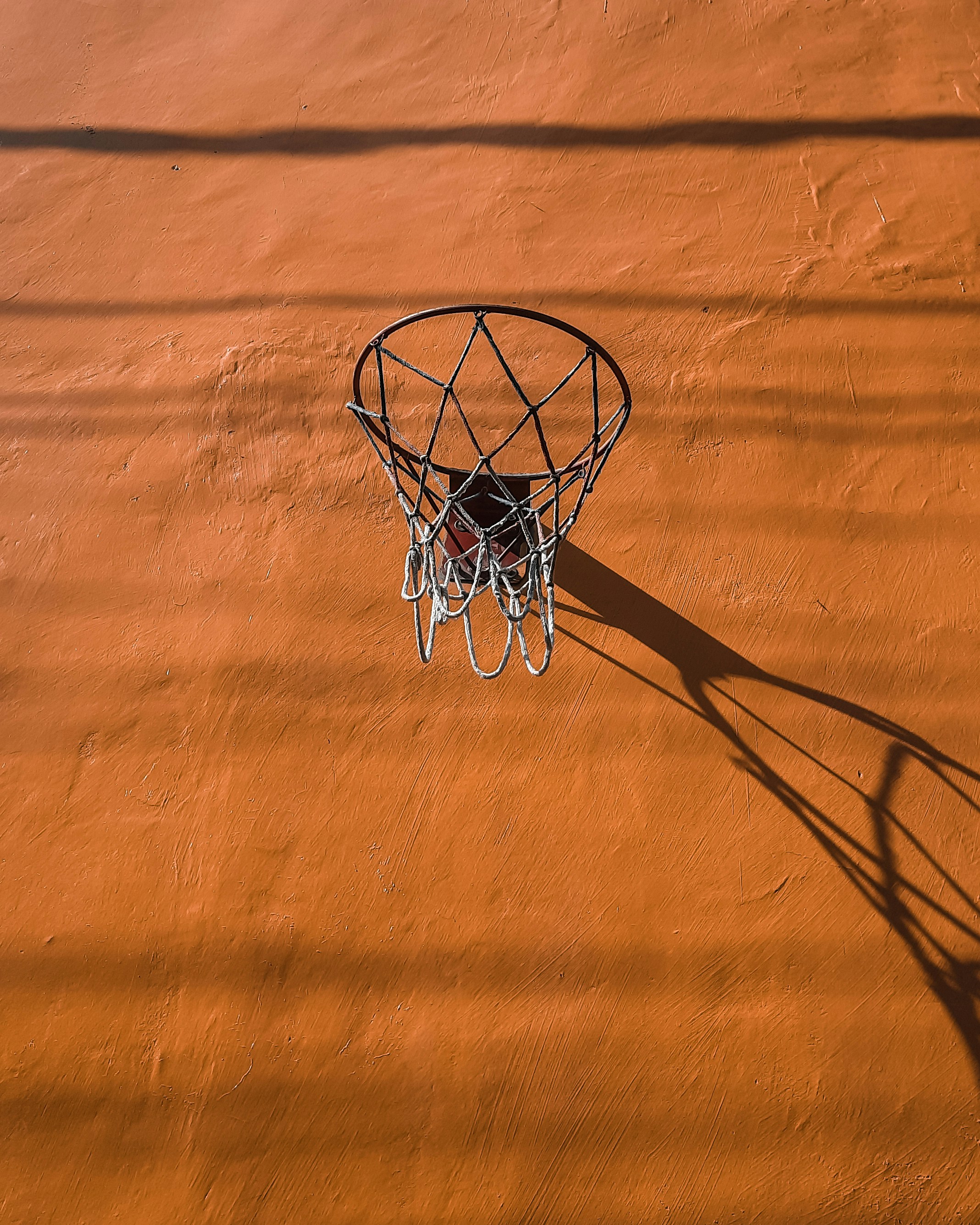 Basketball hoop casting shadows against a vibrant orange wall, highlighting the contrast between sport and stillness.