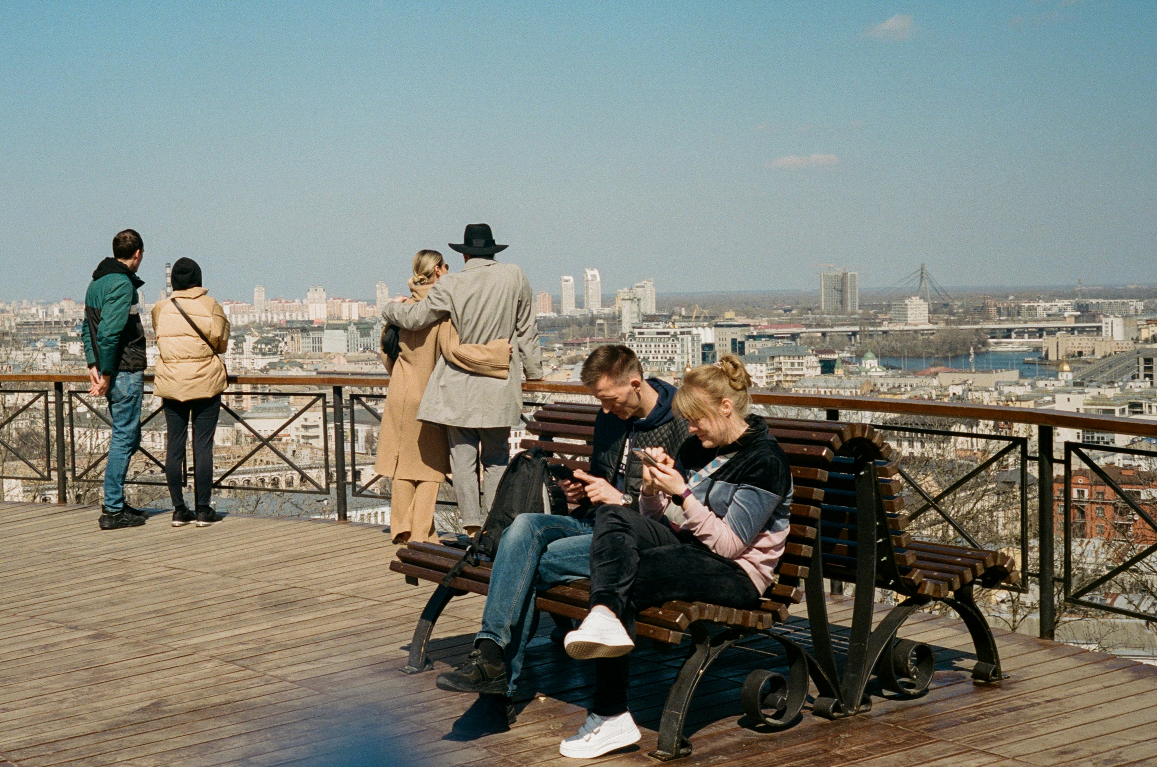Two individuals sit on a bench engrossed in their phones, while couples enjoy the panoramic city view behind them.