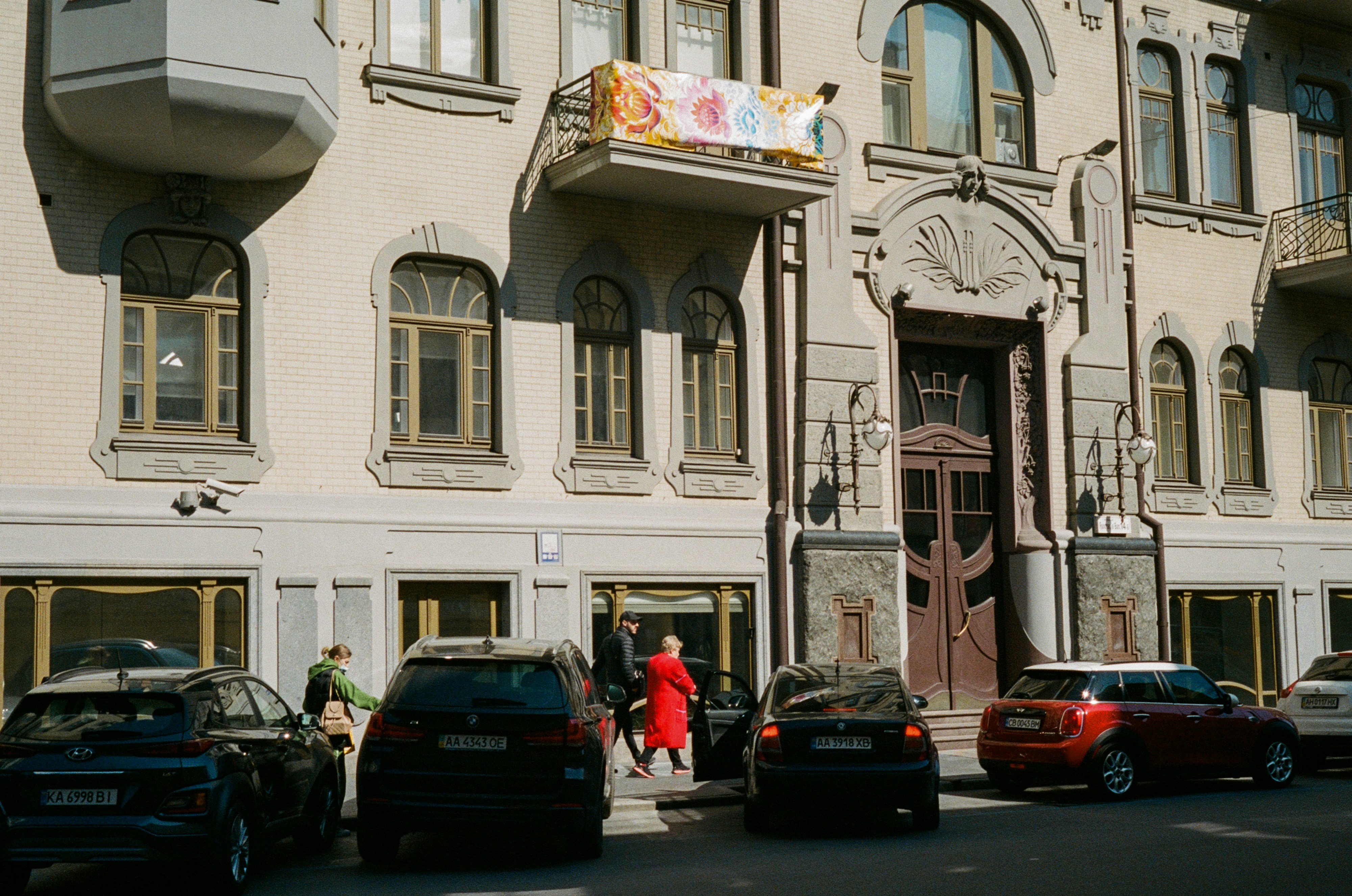 Red-clad pedestrian at ornate building entrance under sunlit row of cars