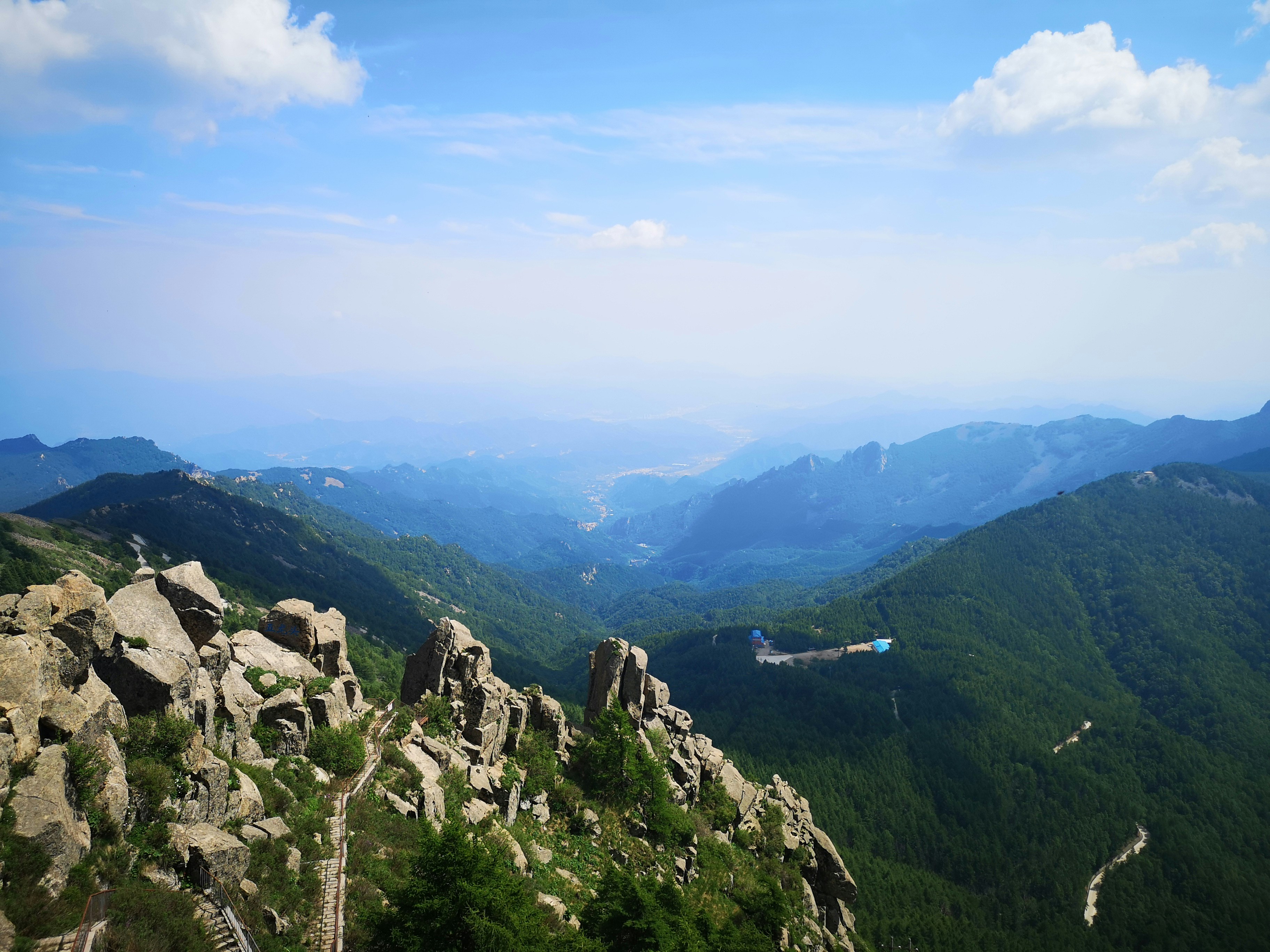 Vast mountain landscape with rocky outcrops and lush greenery under a bright blue sky. A winding path leads through the terrain.