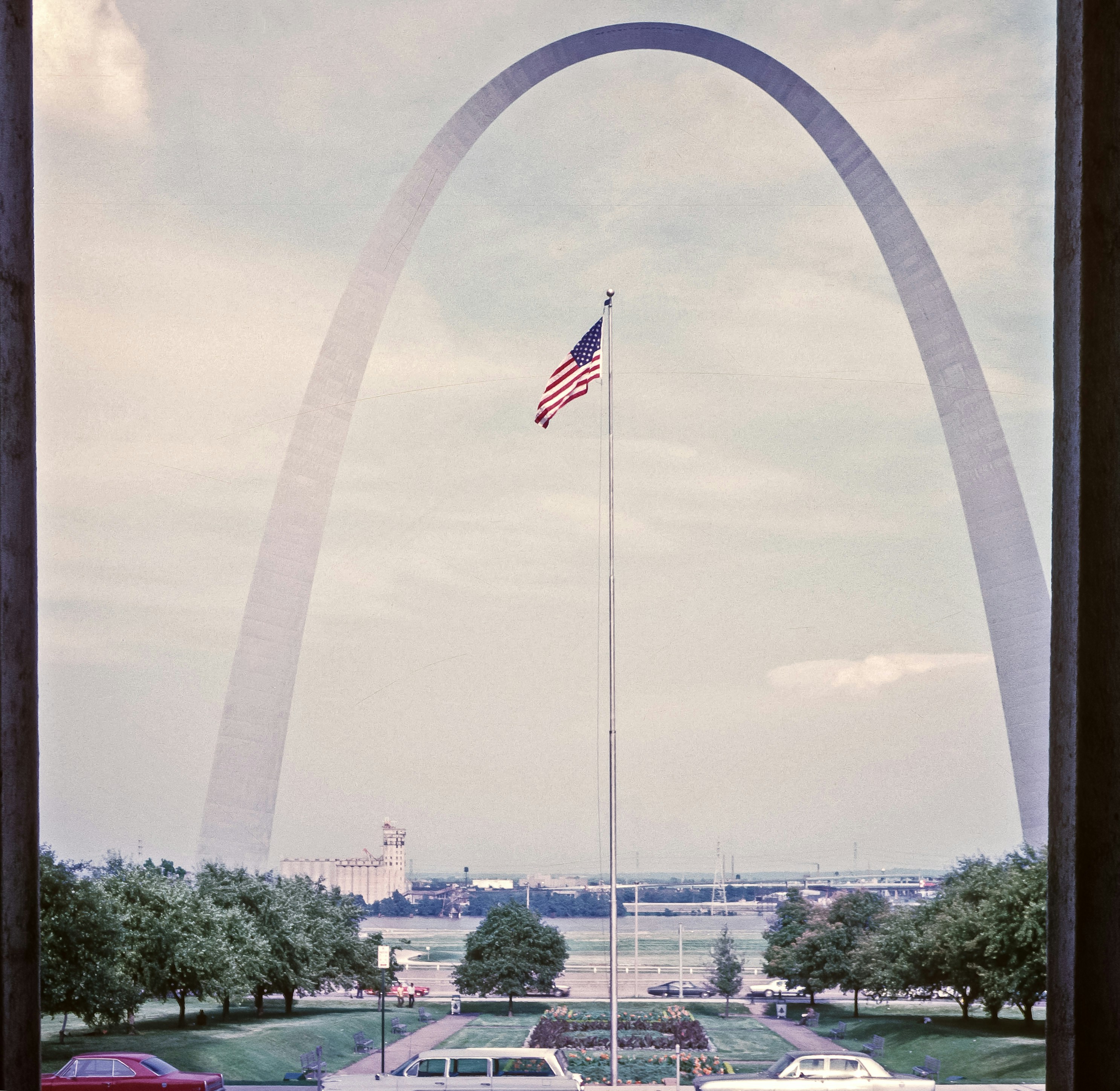 American flag on a tall pole stands in front of the Gateway Arch, with a park, trees, and vehicles in the foreground.